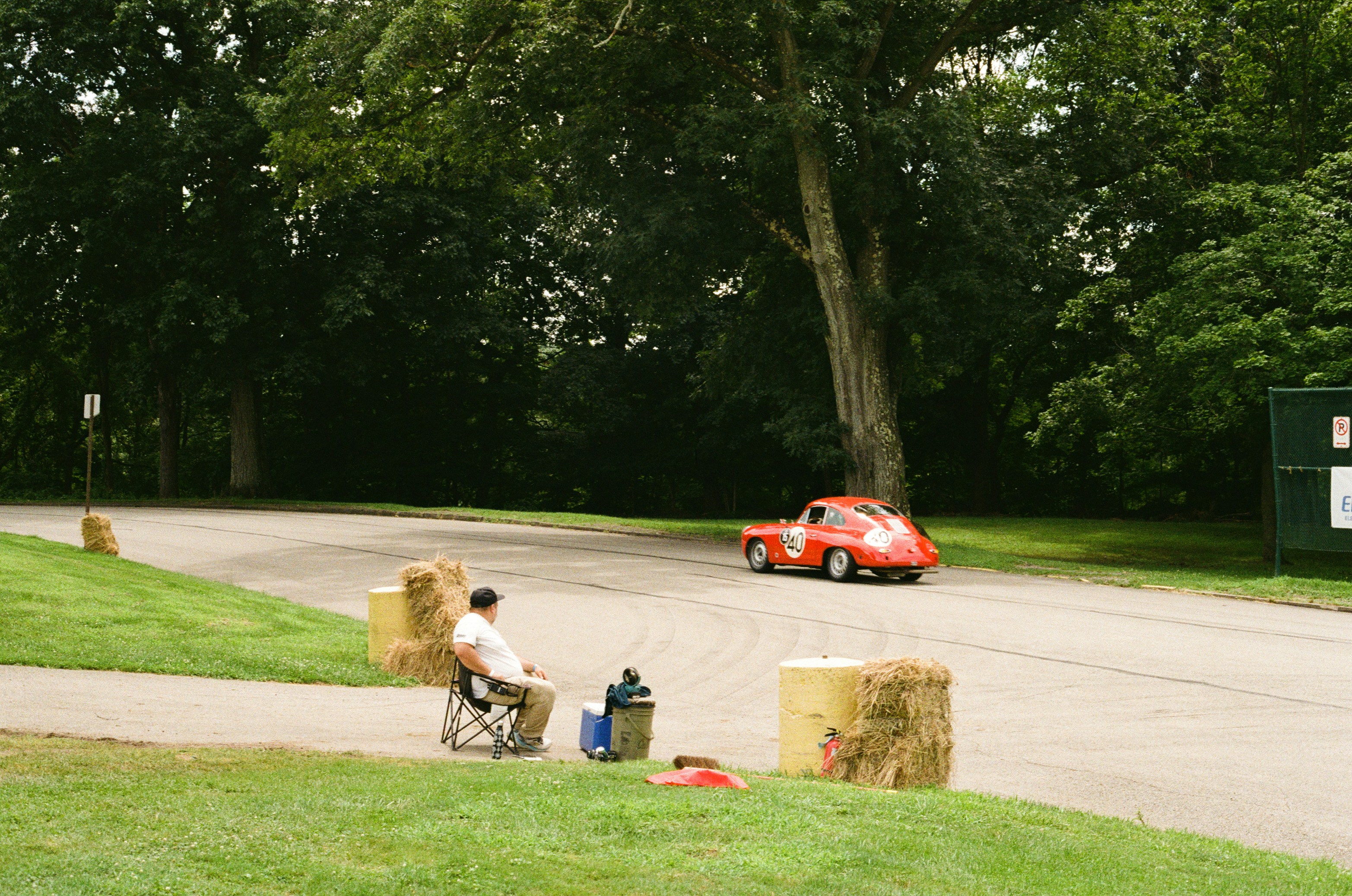 A small red vintage car rounds a curve while a spectator sits on a folding chair by hay bales on a grassy park-like course.