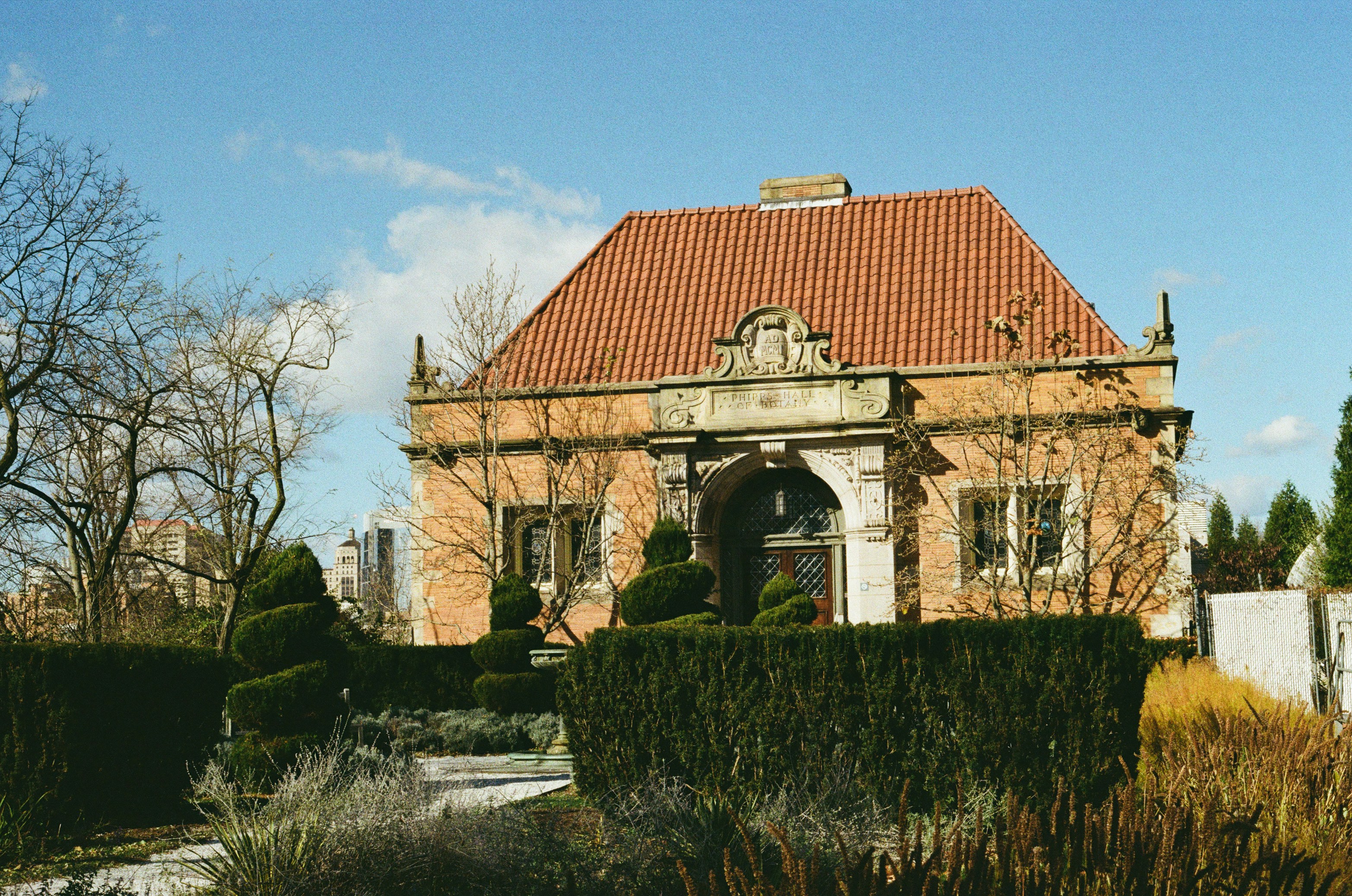 A brick villa with a tiled roof and formal hedges stands under a clear sky; manicured shrubs and a stone archway form the foreground.