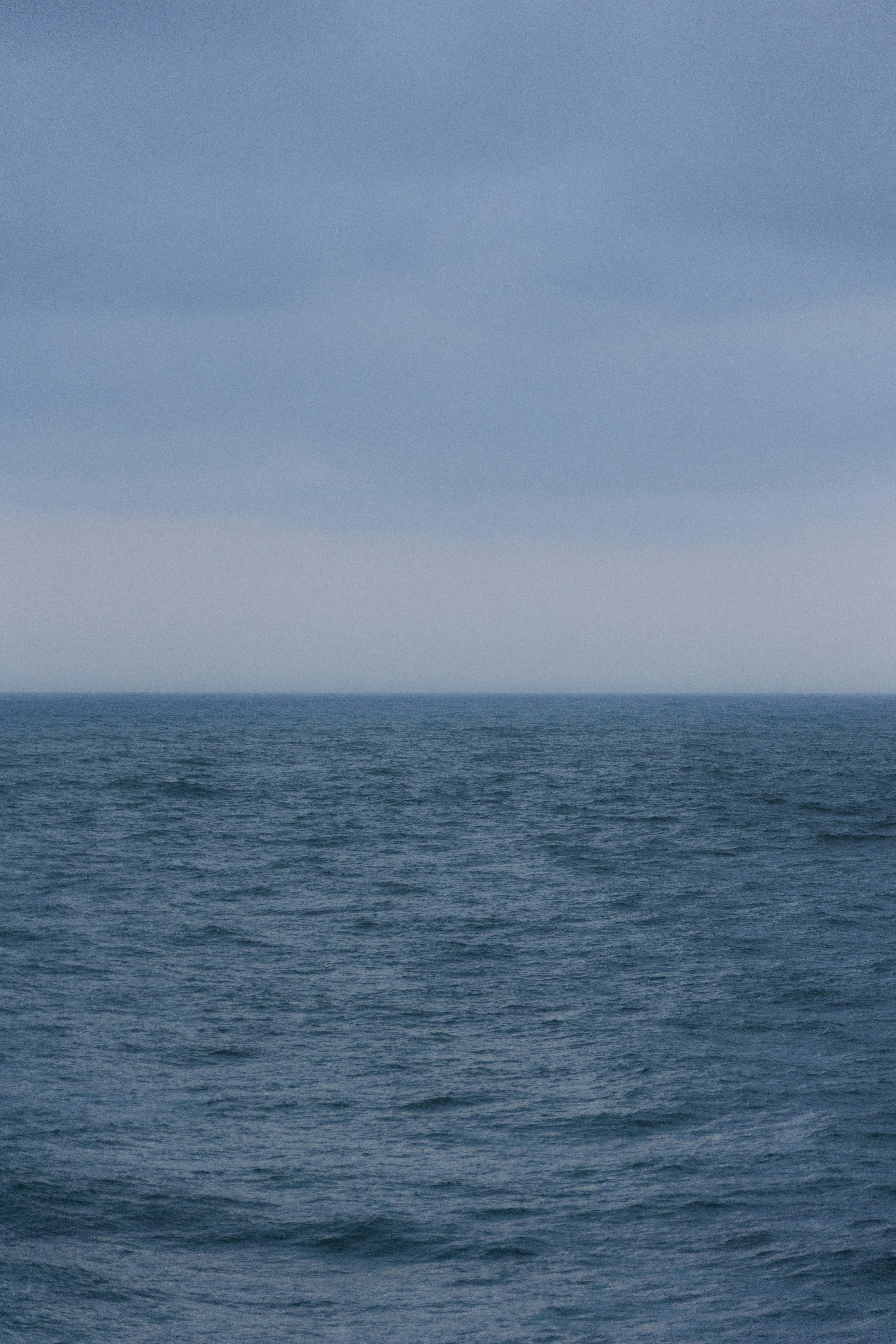 Calm sea under a soft blue sky extends to the horizon, capturing the vast expanse of water with gentle waves and a serene atmosphere.