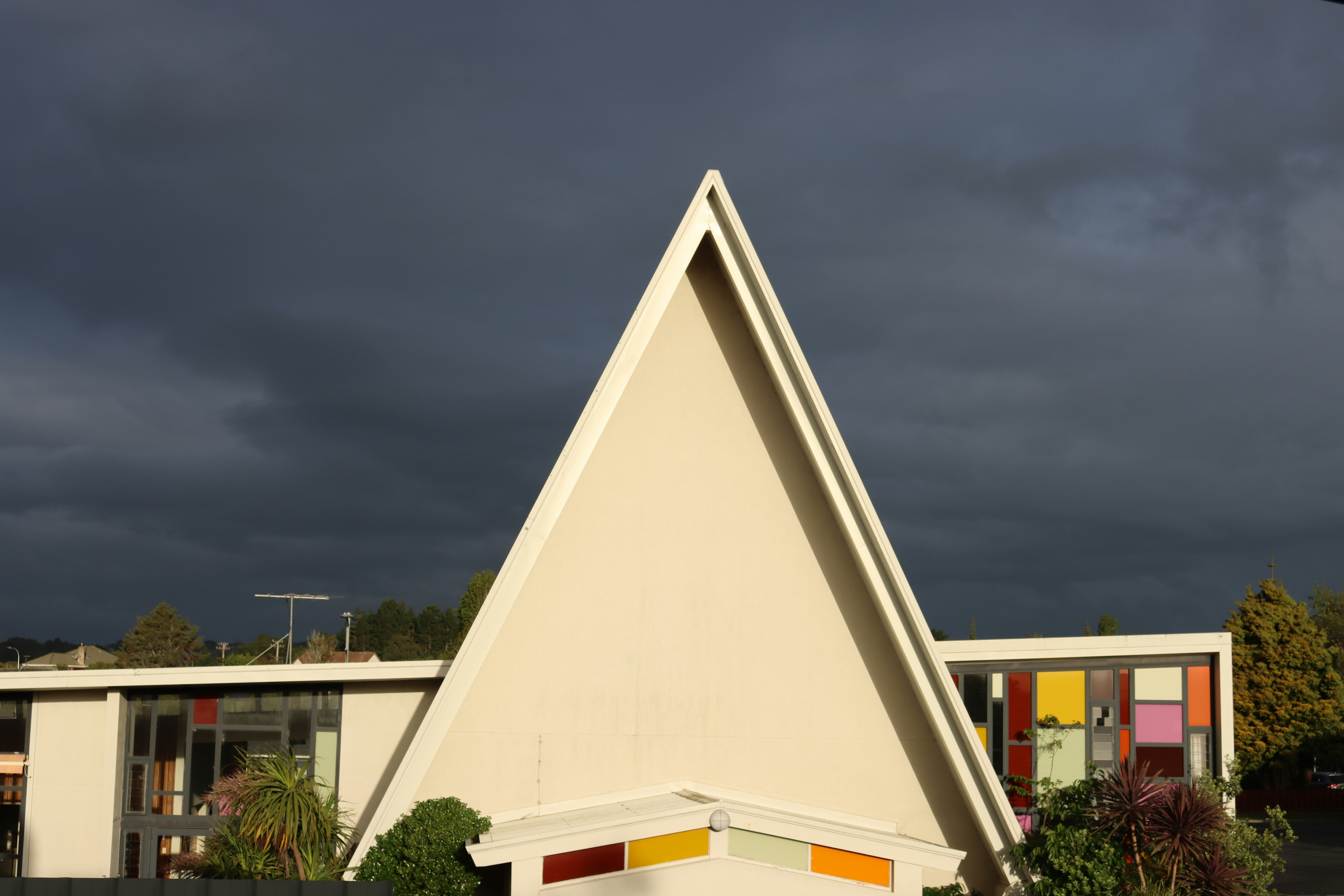 A sharp, triangular rooftop of a modern building rises against a dark, dramatic sky, with colorful storefront windows and surrounding greenery at the base.
