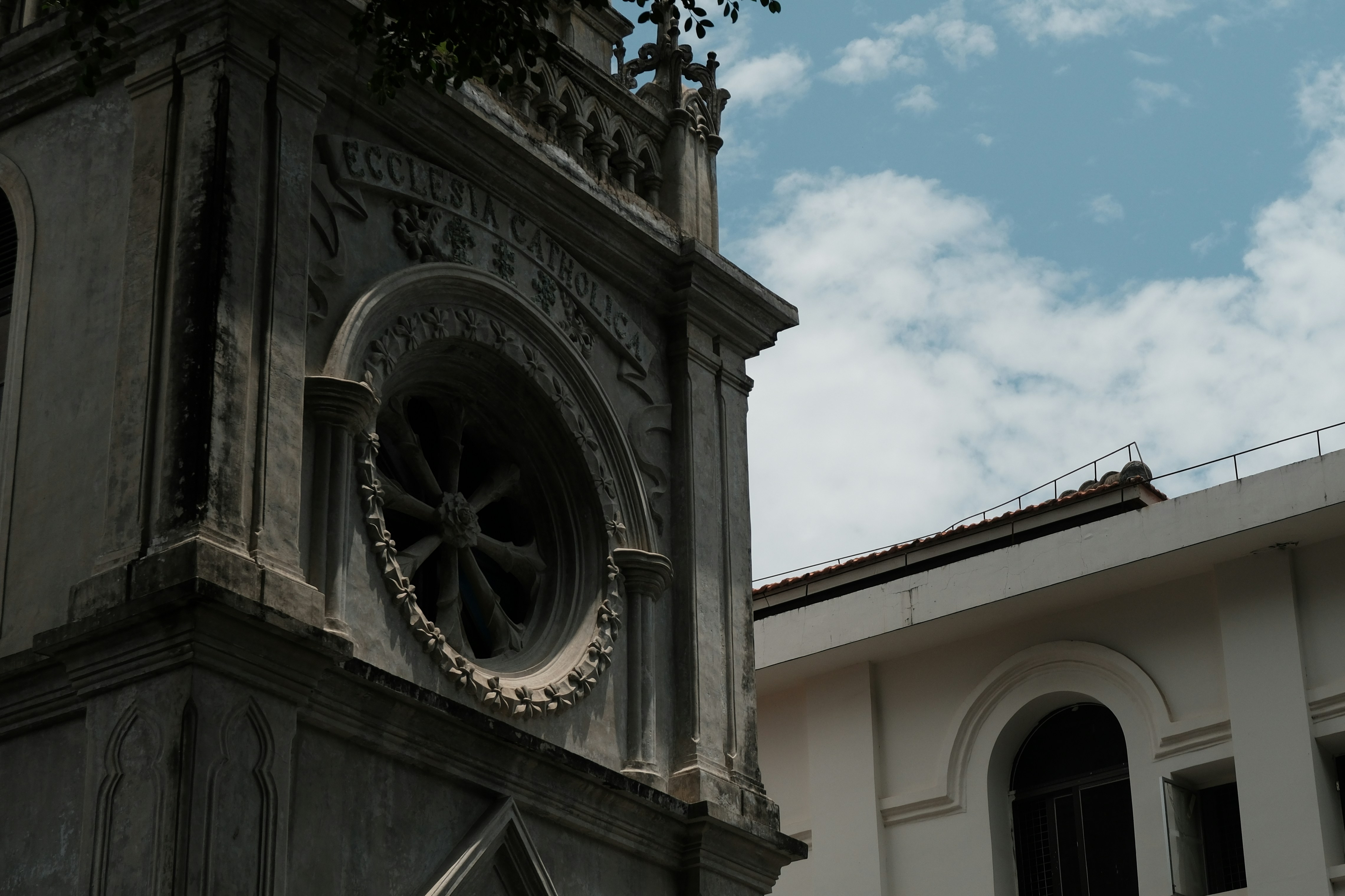 A weathered Catholic church facade with a circular rose window, ornate stone details, and a soft blue sky in the background. The dark shadows and aged architecture give the scene a quiet, historic, and moody atmosphere.