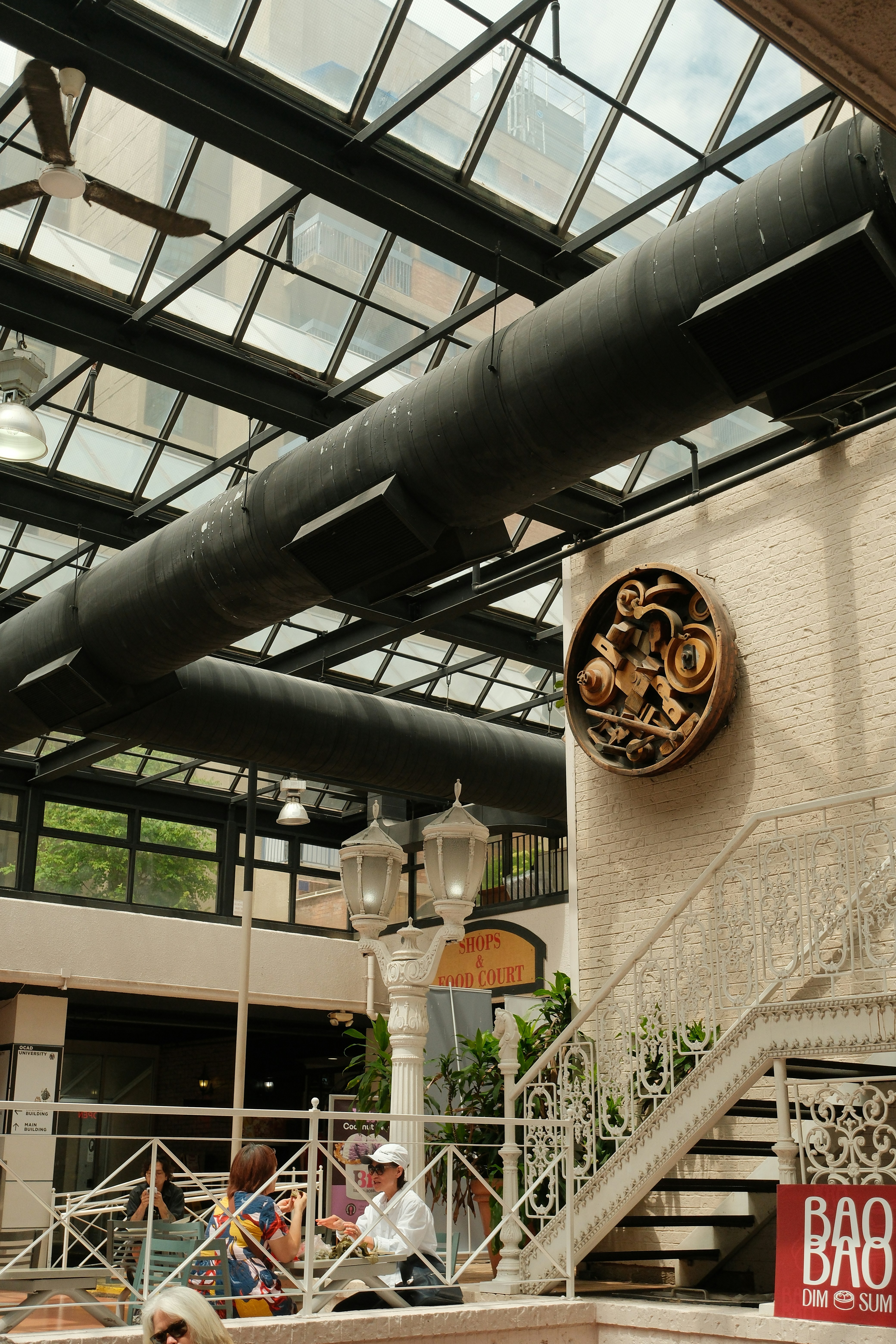 Industrial Atrium: Sunlit Glass Roof and Clockwork Décor Over Café Staircase