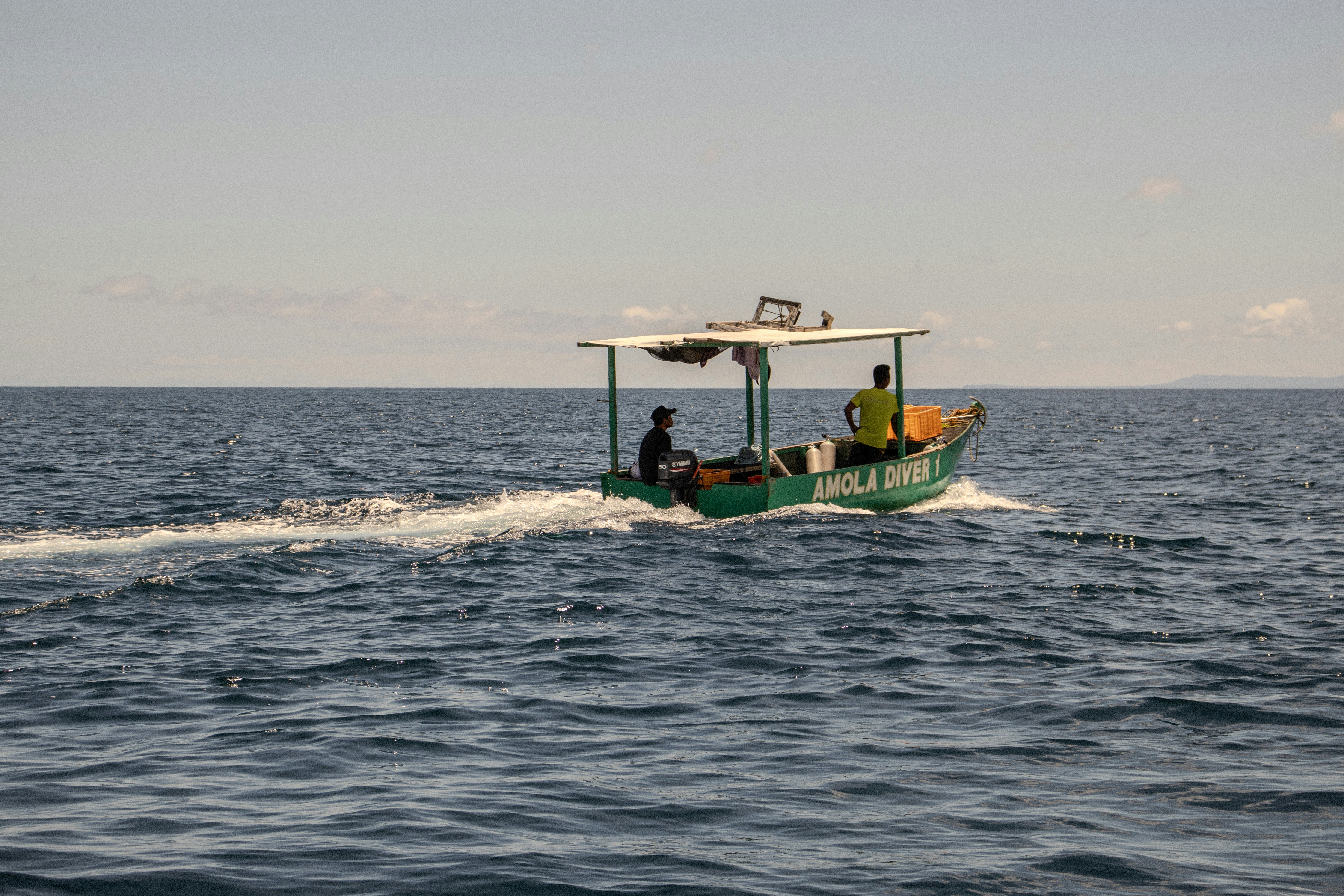 Green diving boat named AMOLA DIVE in the open sea moves across calm water at mid-day sun.