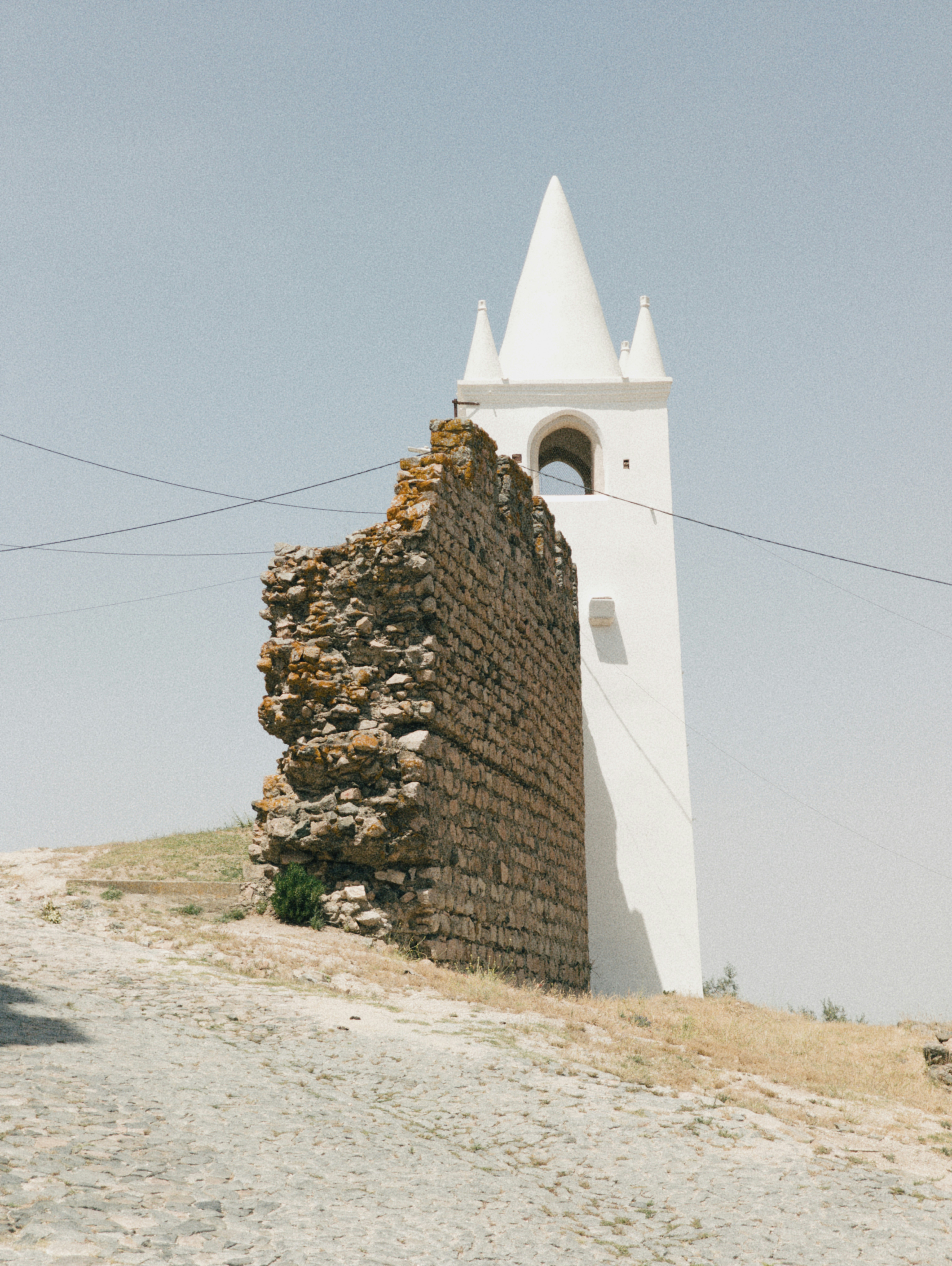 A white church bell tower rises behind an ancient stone wall along a sunlit, dusty path.