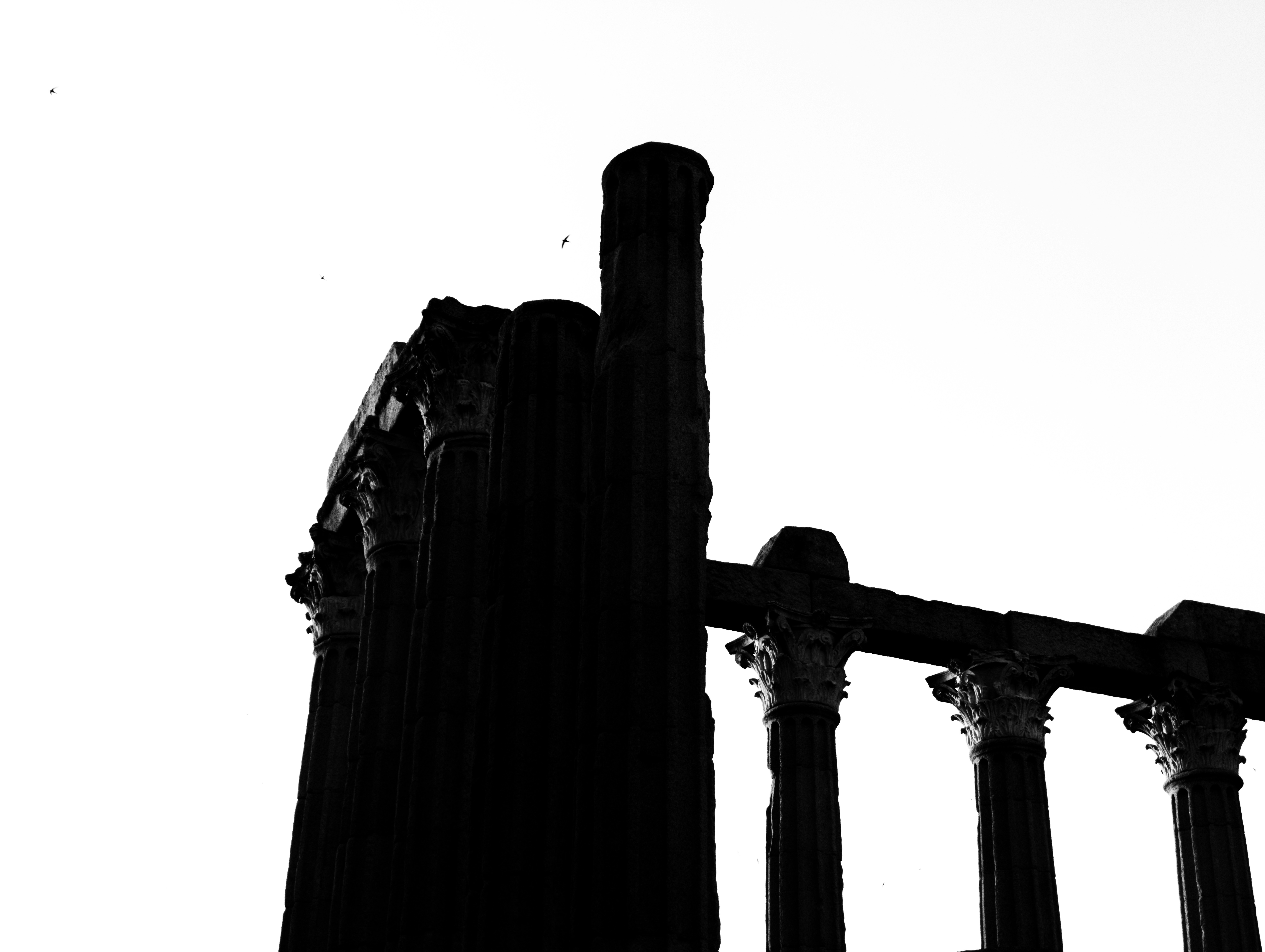 Concrete ruin silhouette with tall columns and a broken arch against a bright sky; monochrome emphasis on architectural decay and negative space.