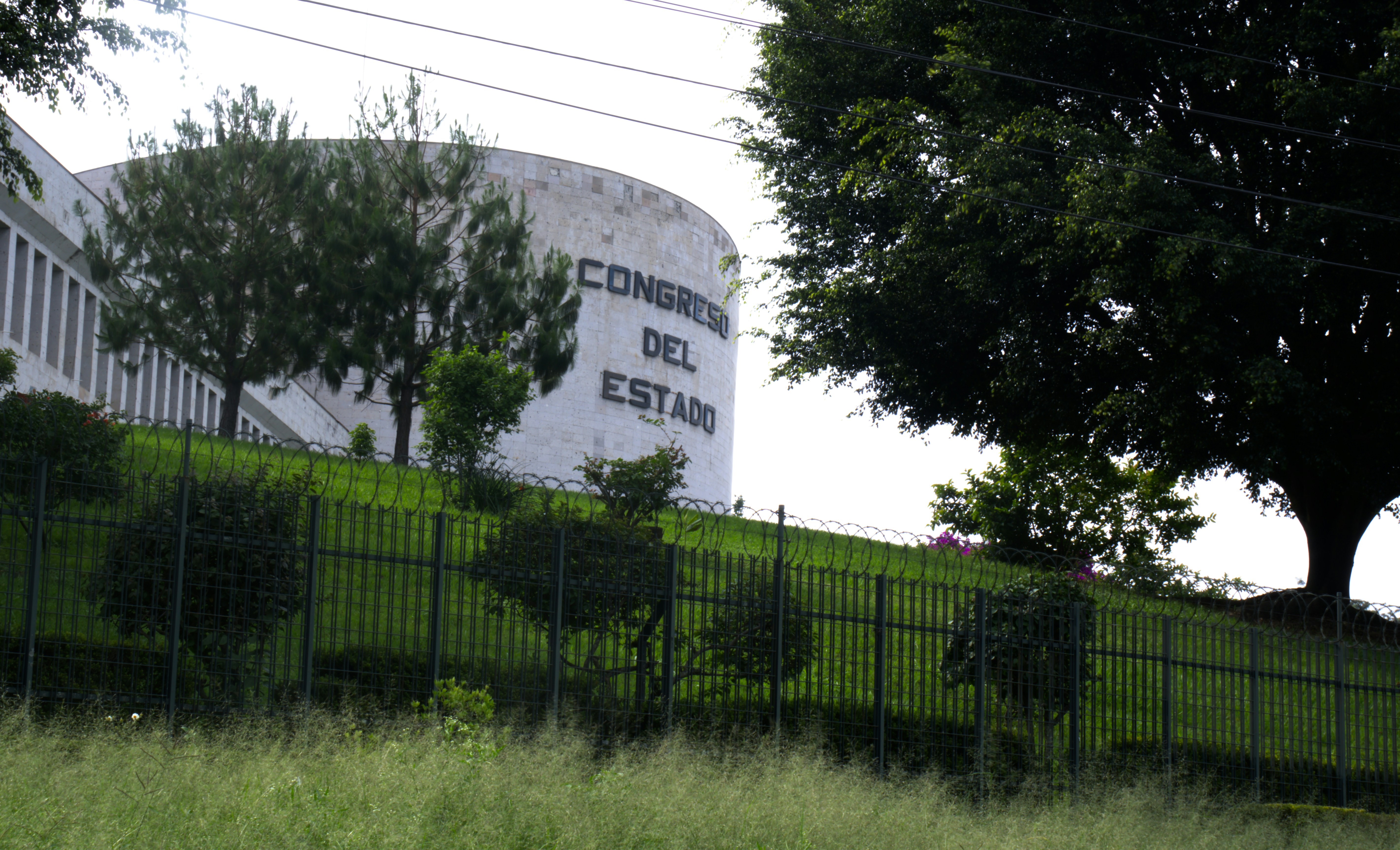 A water tower with 'CONGRESO DEL ESTADO' text sits behind a green metal fence and a grassy foreground, under a tree-lined sky.