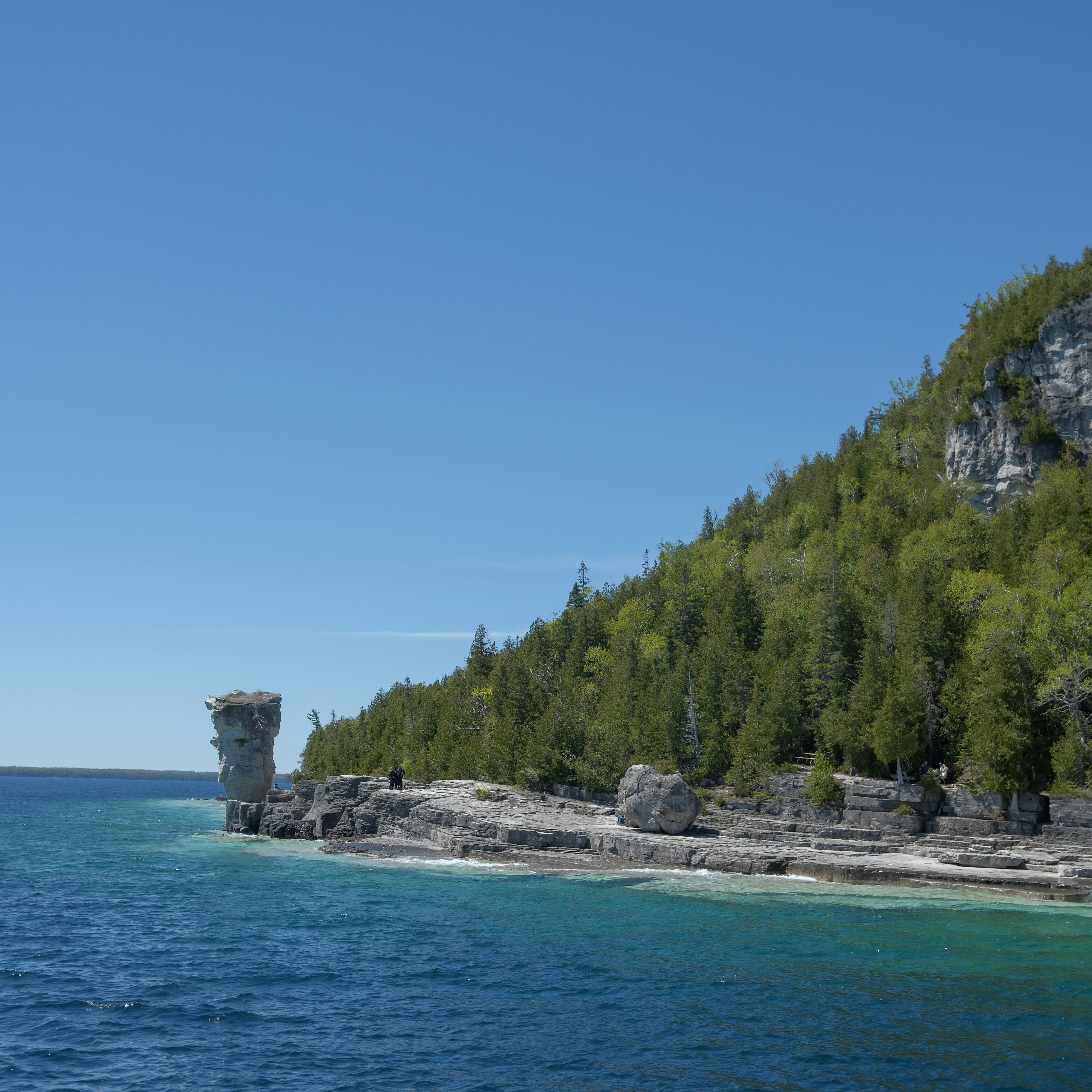 Turquoise sea meets a rugged rocky shoreline backed by a forested cliff; a lone rock formation juts from the water near the left-center, with a calm coastal scene and clear sky.