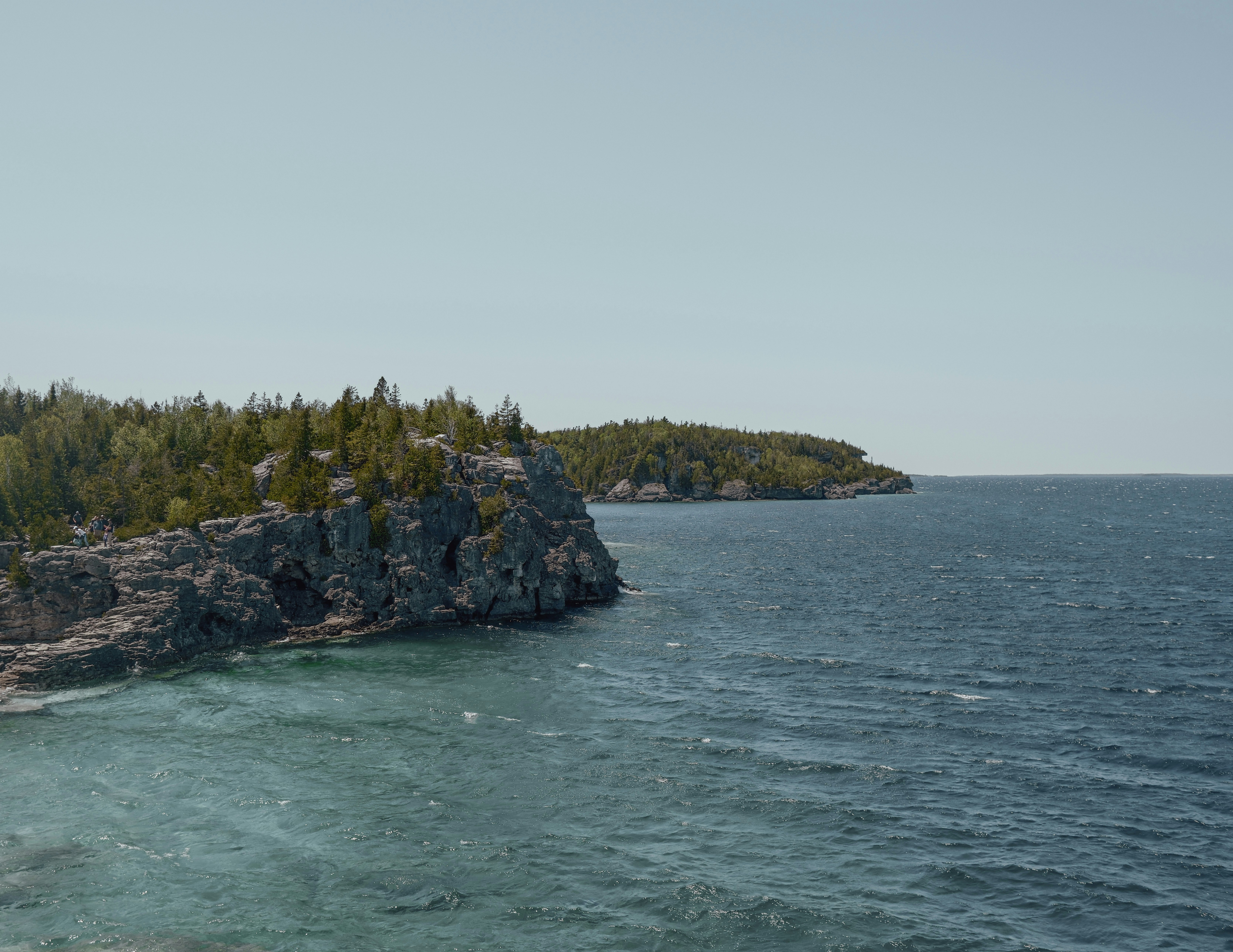 A beautiful wide view of a rugged rocky cliffside drop-off into a vast blue lake. The edge of the land is lined with a thick forest of green evergreen trees, and the water changes into a bright turquoise shade right next to the white stones. It has an open, wild feeling that works perfectly for regional travel guides, hiking maps, or cottage country vacation promotions.