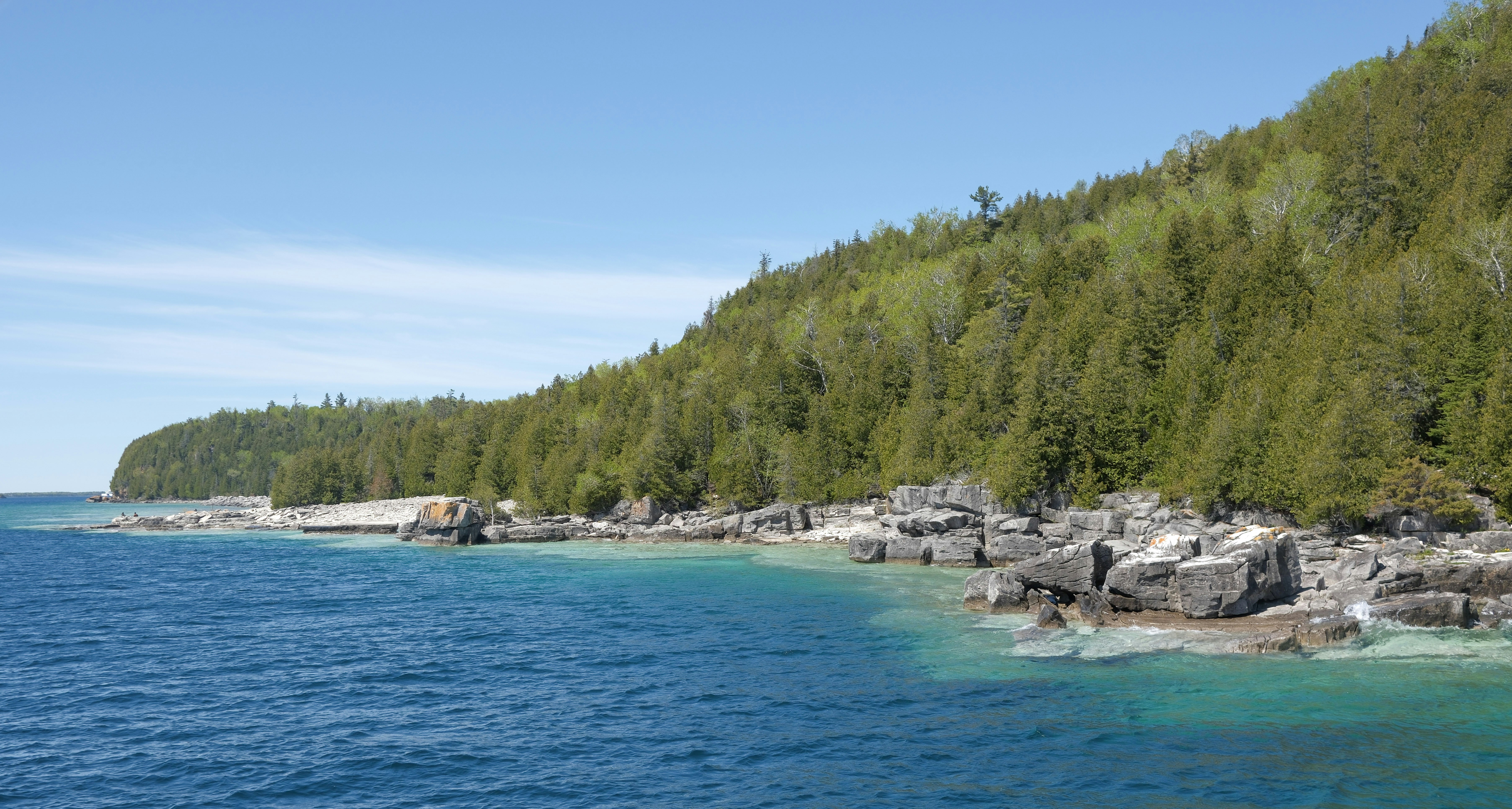 A wide horizontal shot tracking a beautiful rocky shoreline where a dense forest meets the clear lake. The water near the white limestone blocks is a striking emerald green color before turning into a deep navy blue further out. The sun is shining bright, making all the tree leaves stand out cleanly against the rock layers. This raw file is an excellent visual asset for outdoor gear ads, conservation websites, or travel diaries.