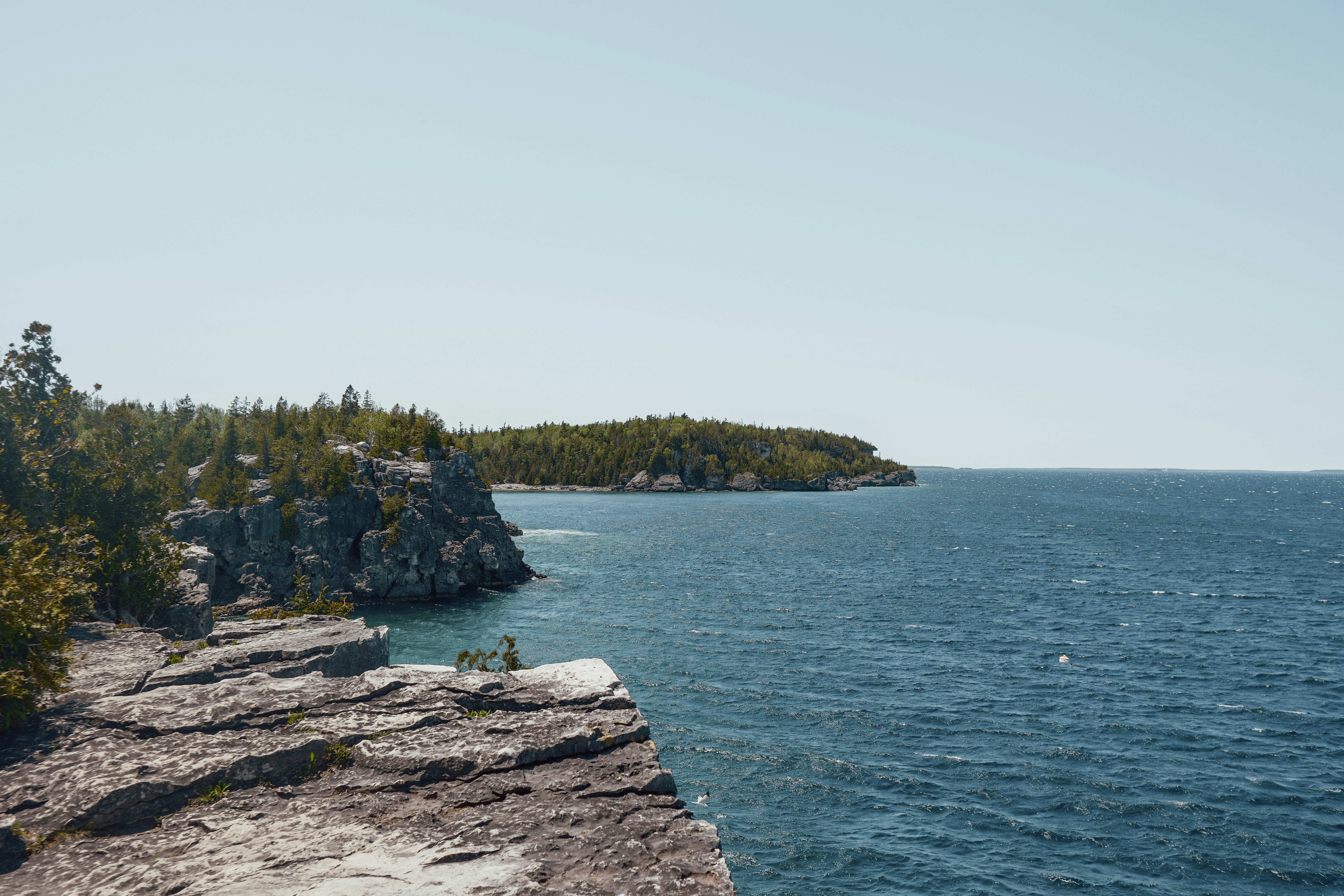 A dramatic landscape shot taken from the top of a high cliff looking out across a massive body of water. The grey, weathered rocks in the foreground drop off into a deep blue lake that stretches all the way to a distant forested peninsula. The clear sky and bright sun make the entire scene feel huge and open. This premium raw file is ready to use for travel agencies, destination booking websites, or local guidebooks.