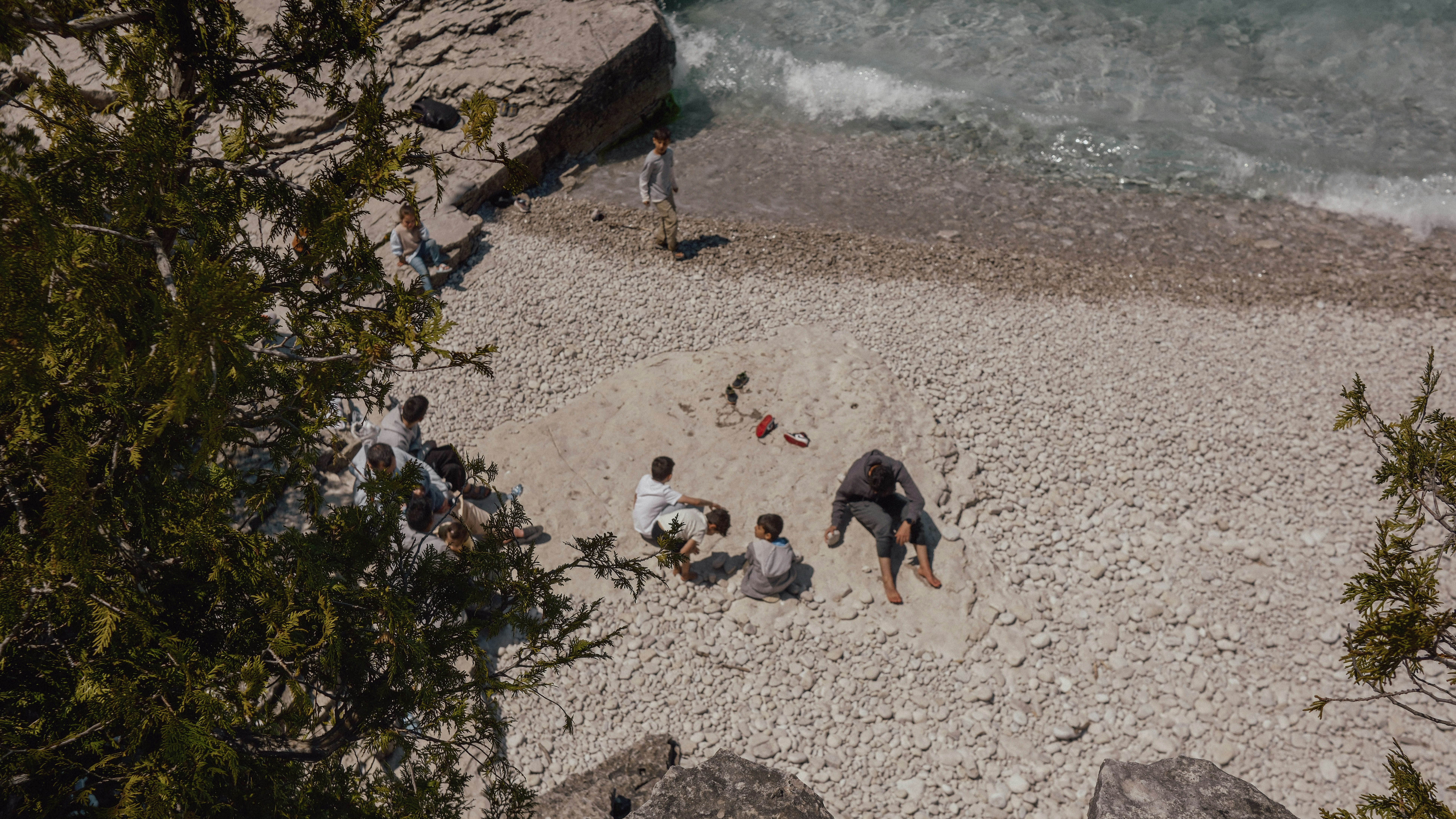 A top-down view looking down at a beautiful white pebble beach filled with families and travelers relaxing in the sun. Some people are sitting together on a massive flat rock slab, while others are walking along the water's edge where clear turquoise waves roll in. A green tree branch frames the left side softly. This lively, unpolished photo shows the authentic community lifestyle of a summer park holiday, perfect for tourism networks or booking websites.