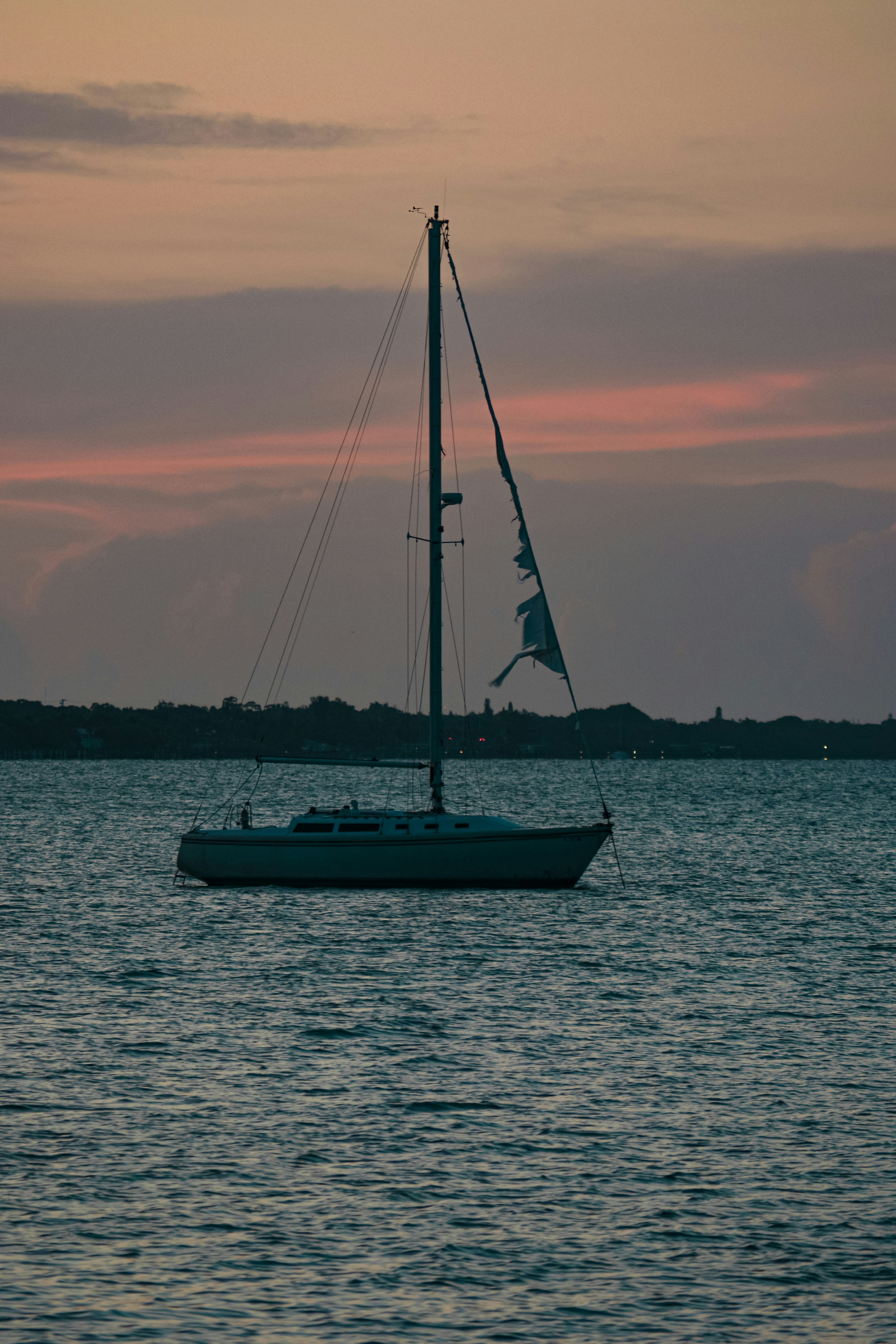 Silhouette of a lone sailboat on calm water at dusk, with a pastel sky and distant shoreline in the background.