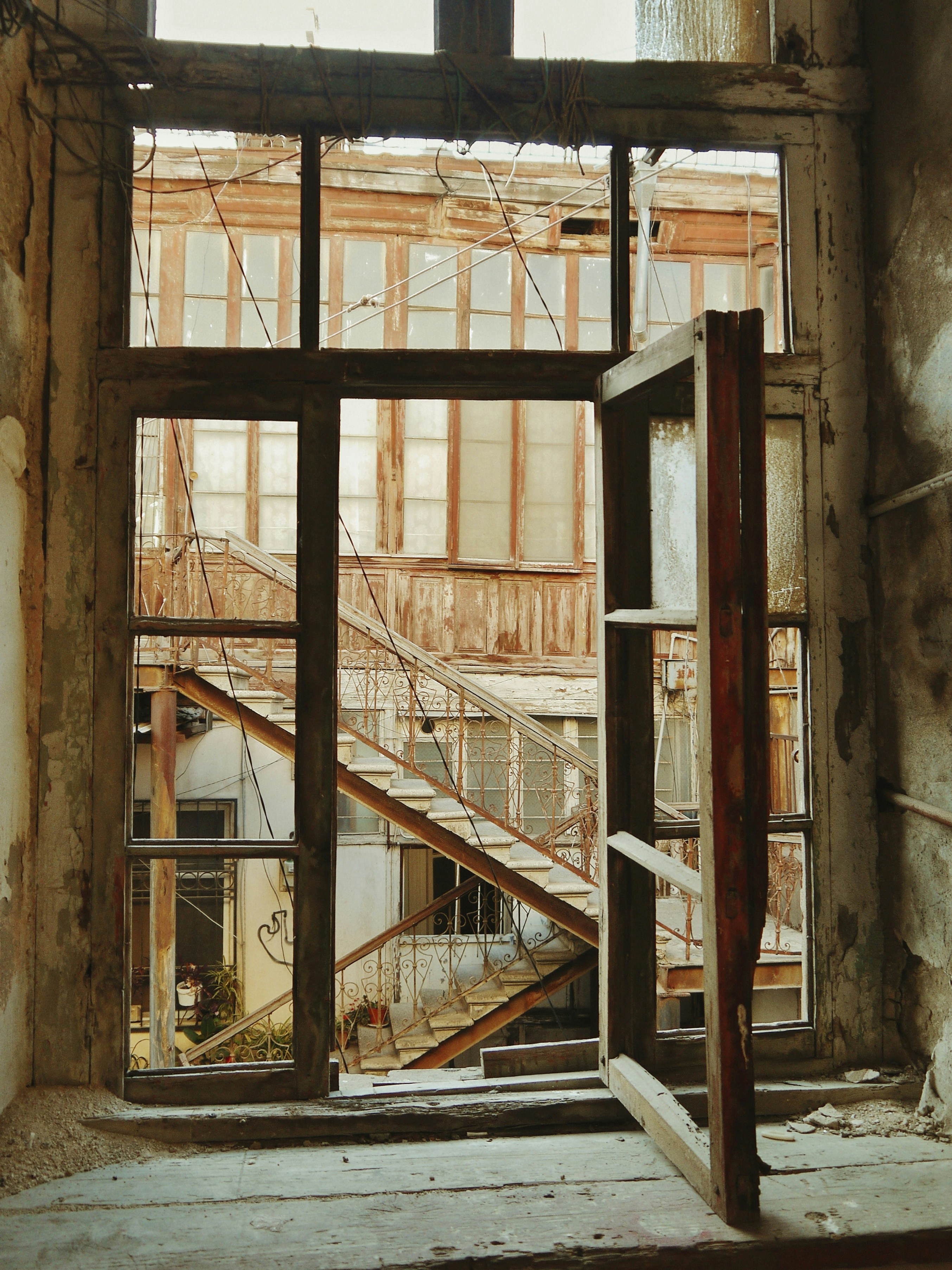 Worn interior with an open window framing a dilapidated outdoor stairwell and brick facade; muted warm tones emphasize textures of wood, metal, and plaster.