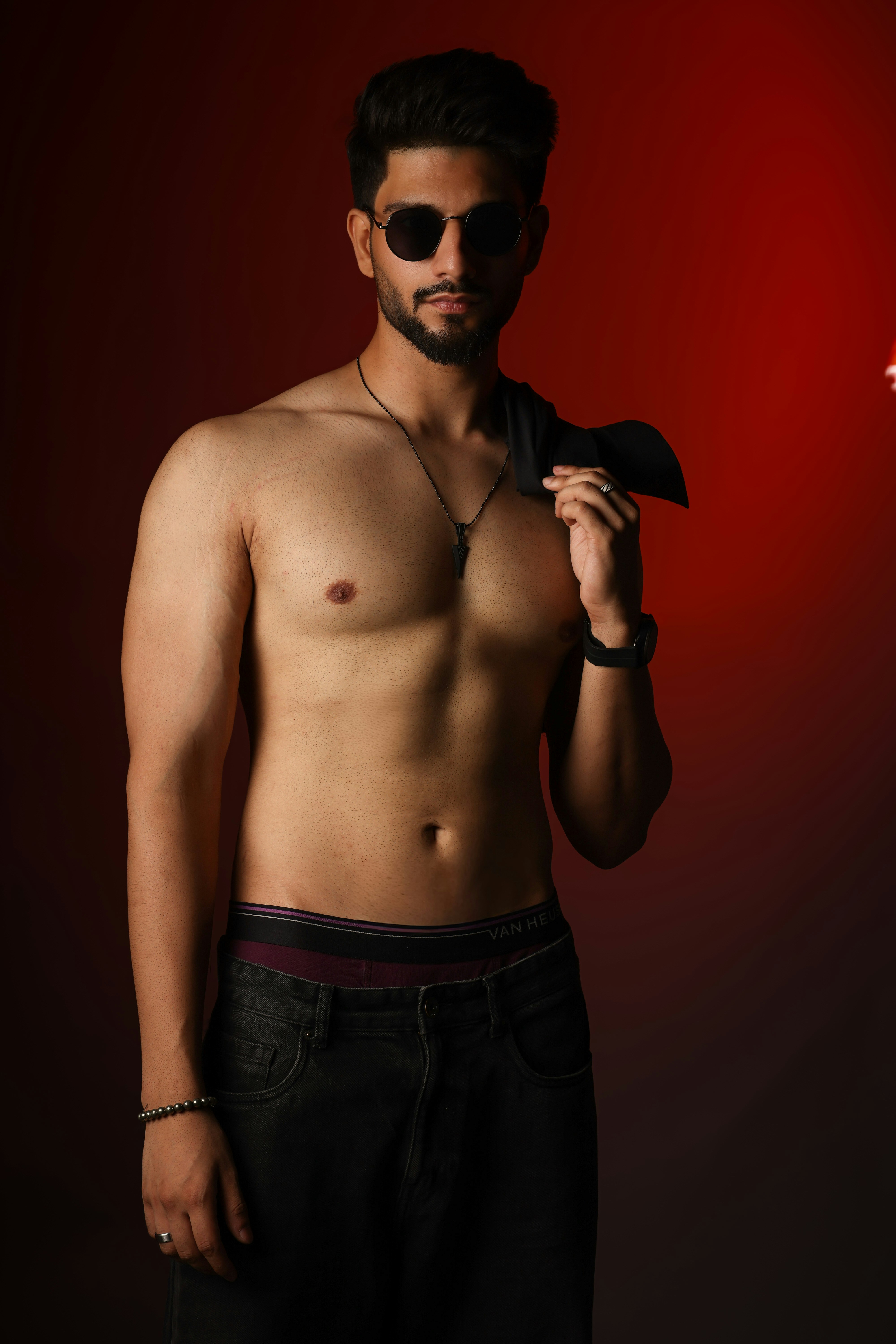 Athletic male model shirtless in a dark studio with a red backdrop and sunglasses, holding a jacket over one shoulder; moody lighting emphasizes defined musculature and sculpted facial features.