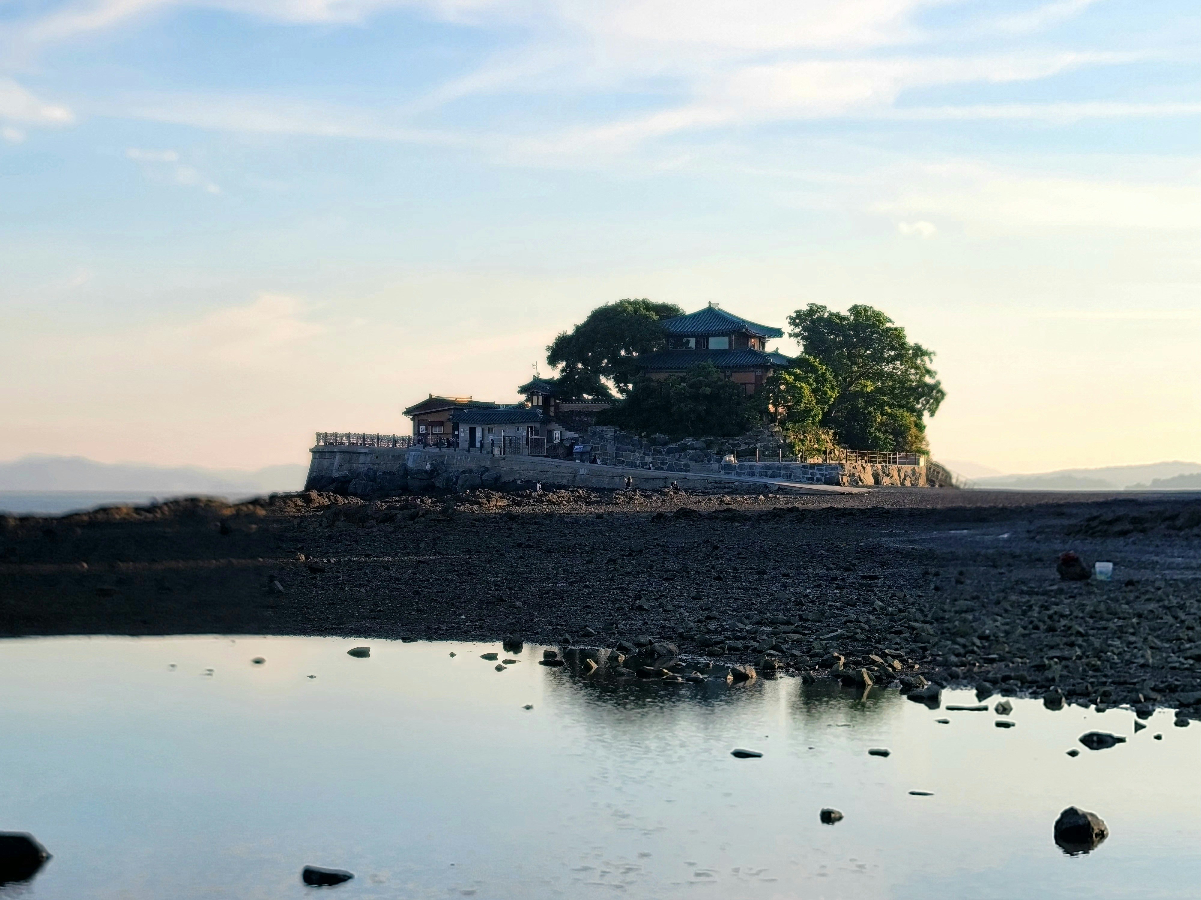 Small island fortress with trees atop a stone wall sits in calm tidal waters; distant shore and rocky shoreline frame a tranquil harbor scene.