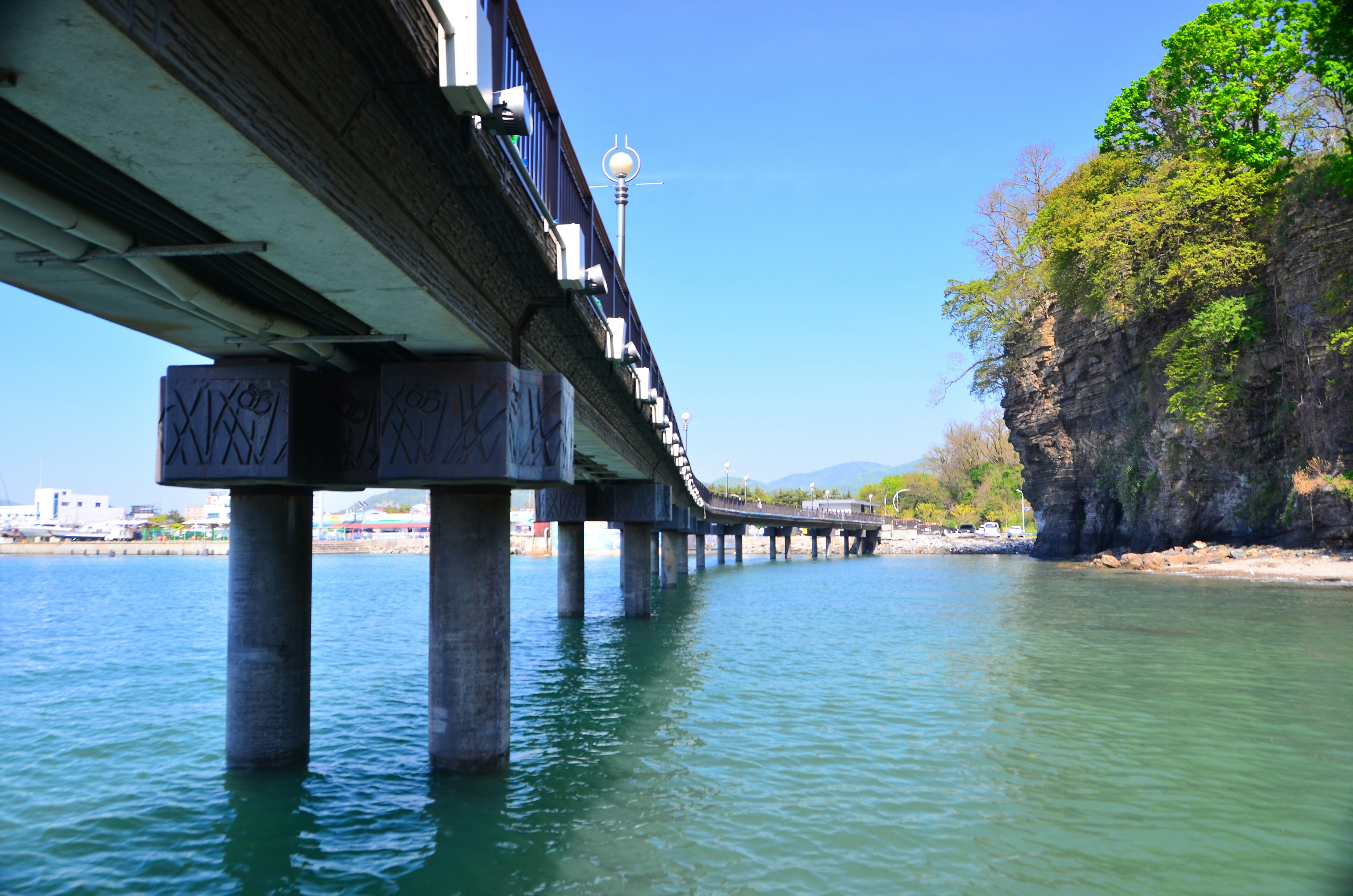 Sunlit bridge spans turquoise water with a rocky cliff and trees in the background, forming a clean, linear composition.