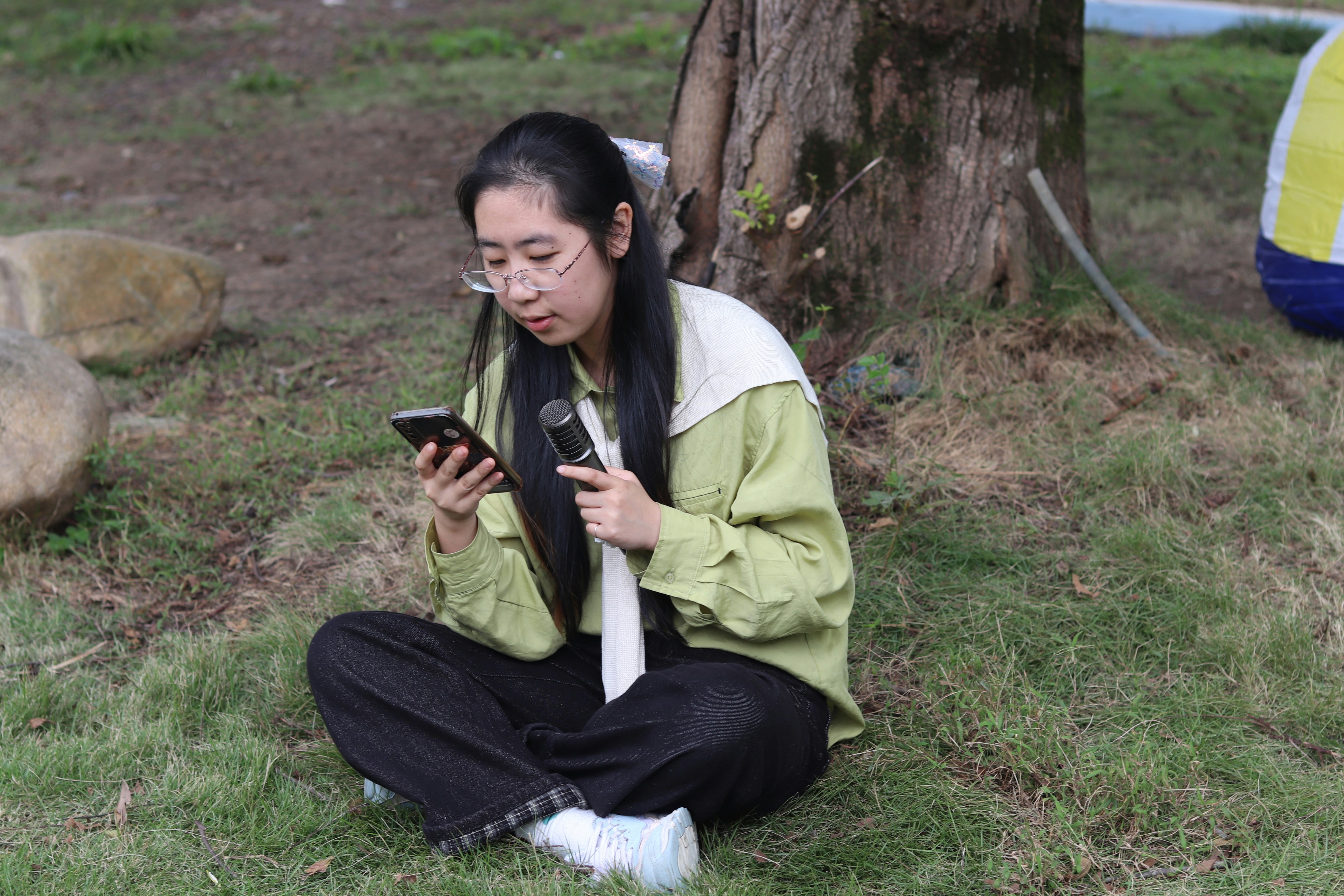 Young woman sitting on grass under a tree, holding a microphone and looking at a phone; appears to be at a casual outdoor event or campsite, with a small tent visible in the background.