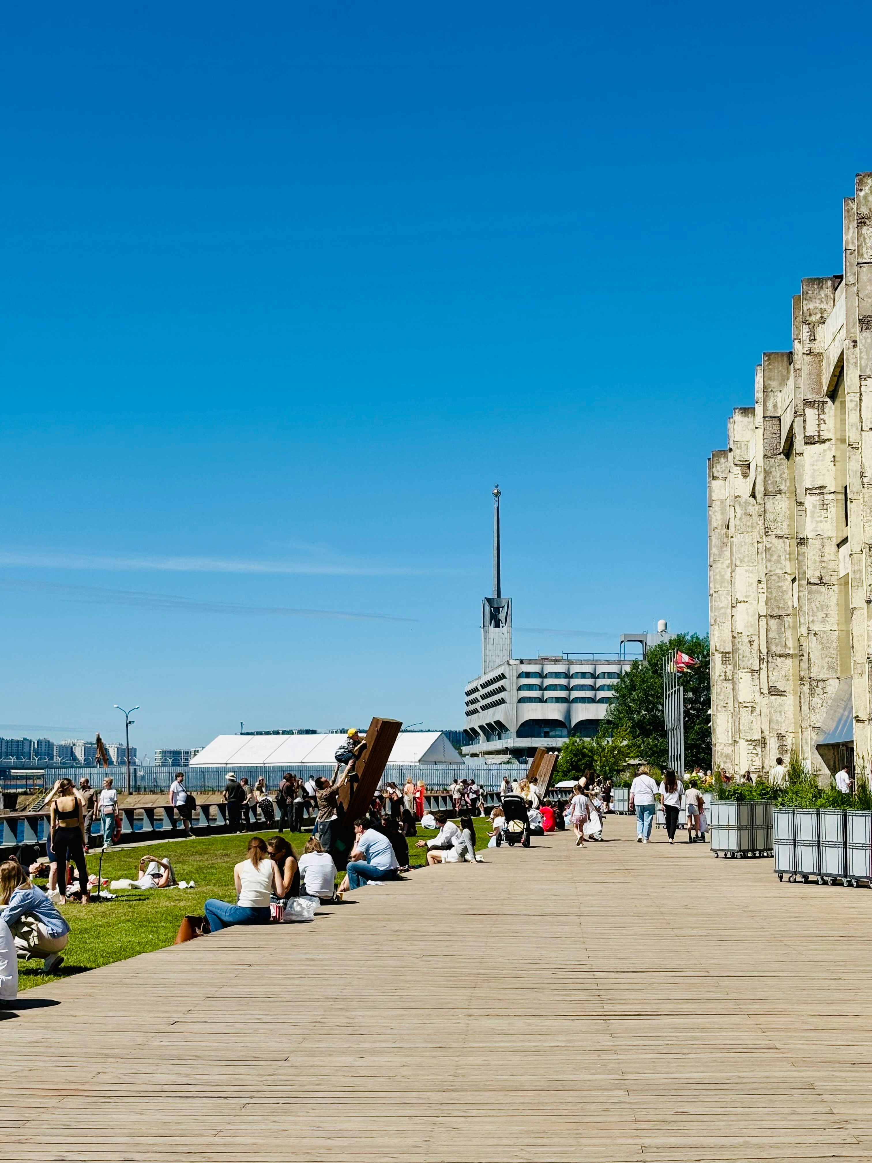People at a sunlit riverside promenade beside a towering modern façade
