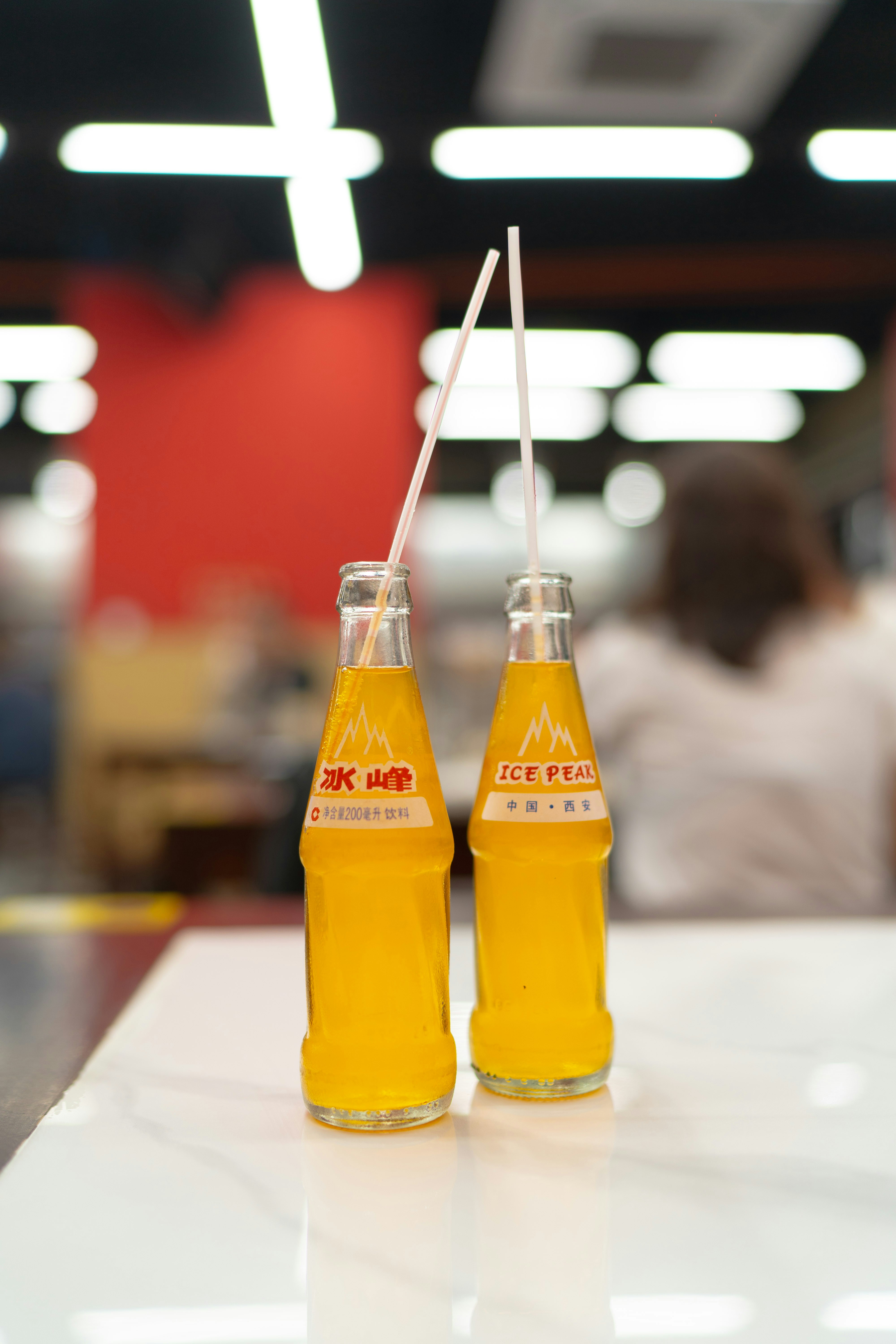 Two glass bottles of orange soda sit on a white counter in a softly blurred cafe scene
