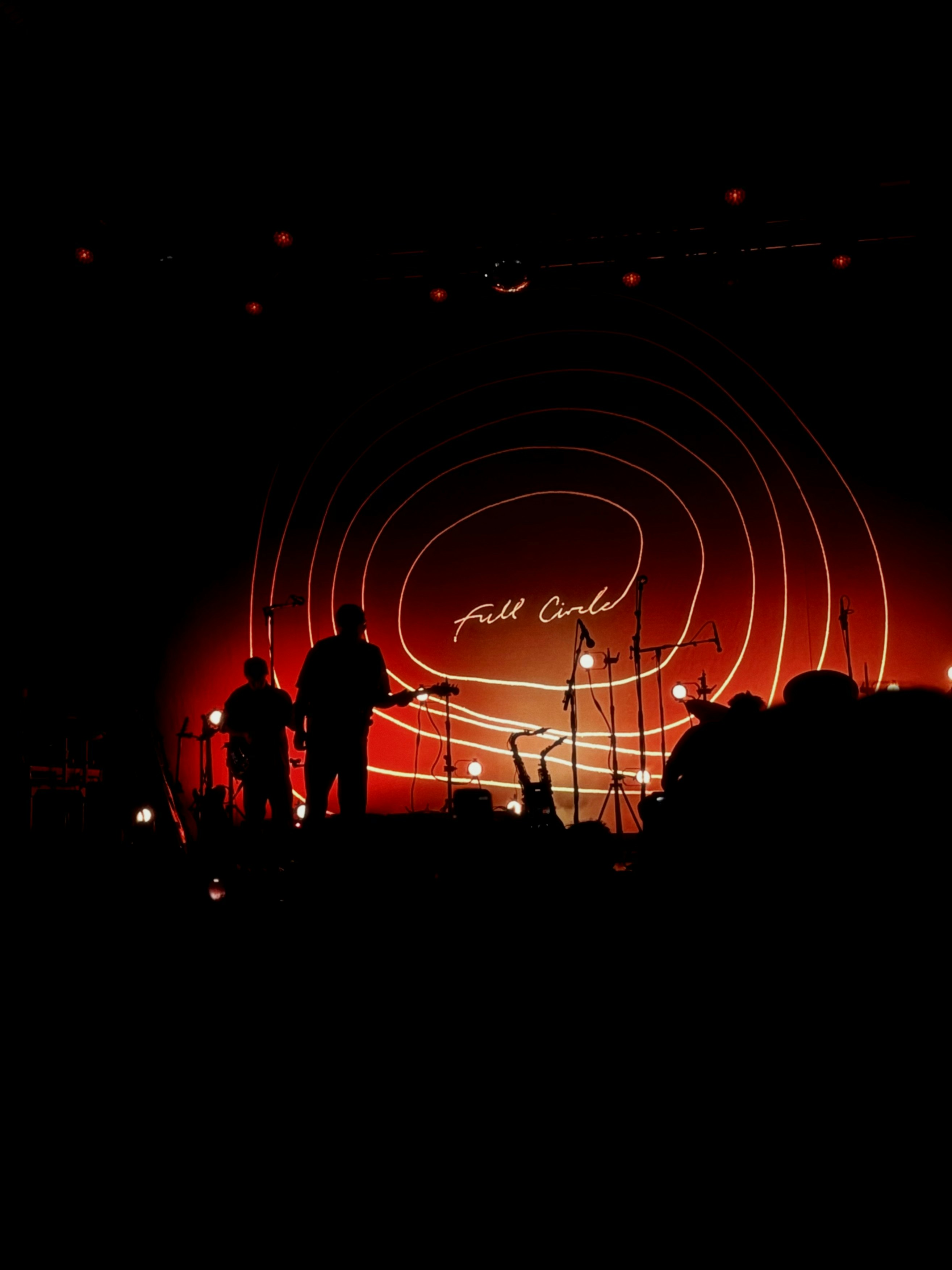 Silhouettes of a live band perform against a red, circular neon backdrop, with spotlighted stage gear and audience hints in darkness.