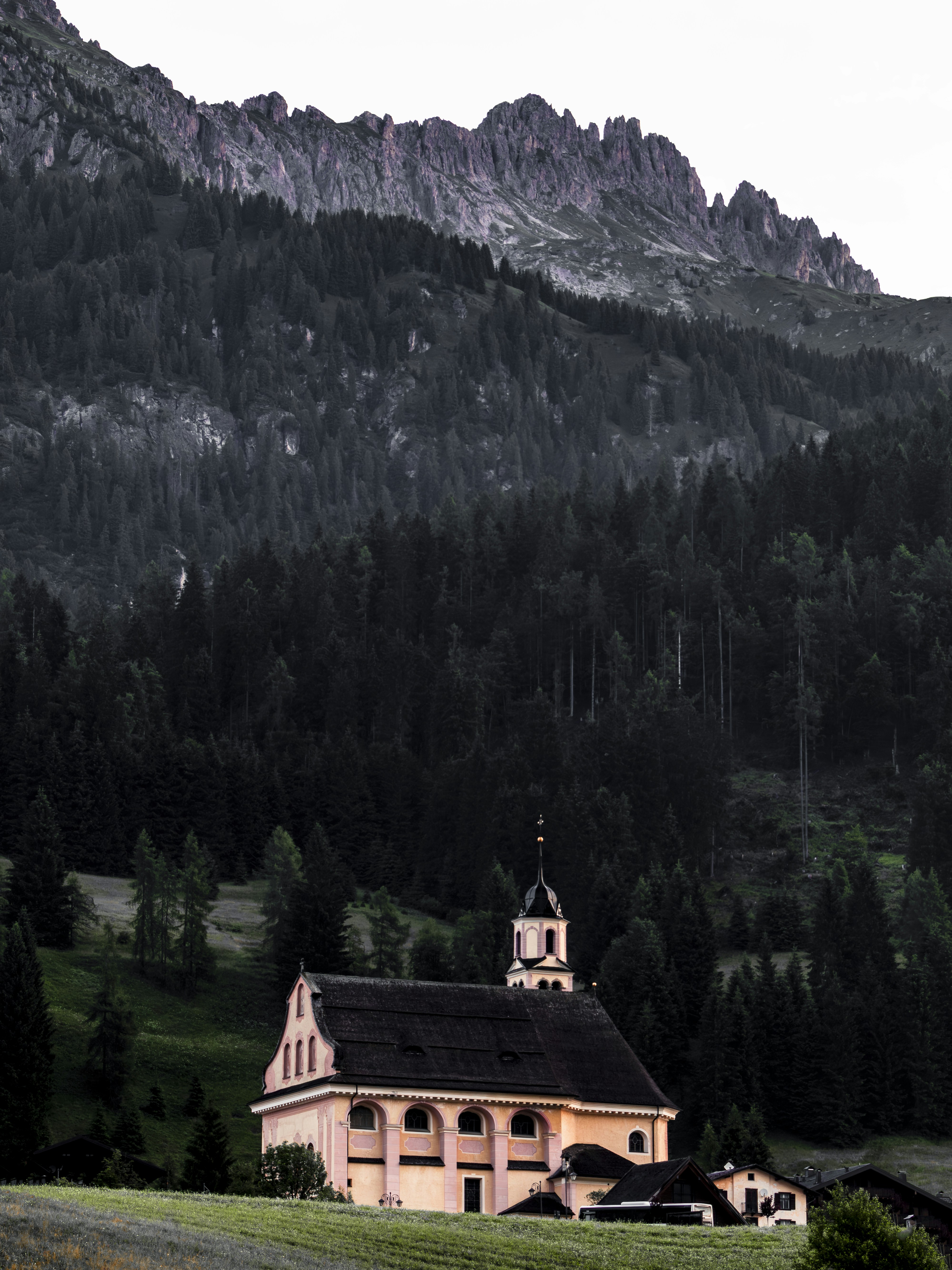Pink church nestled at the foothills with dense pine forest and towering mountains in the background.