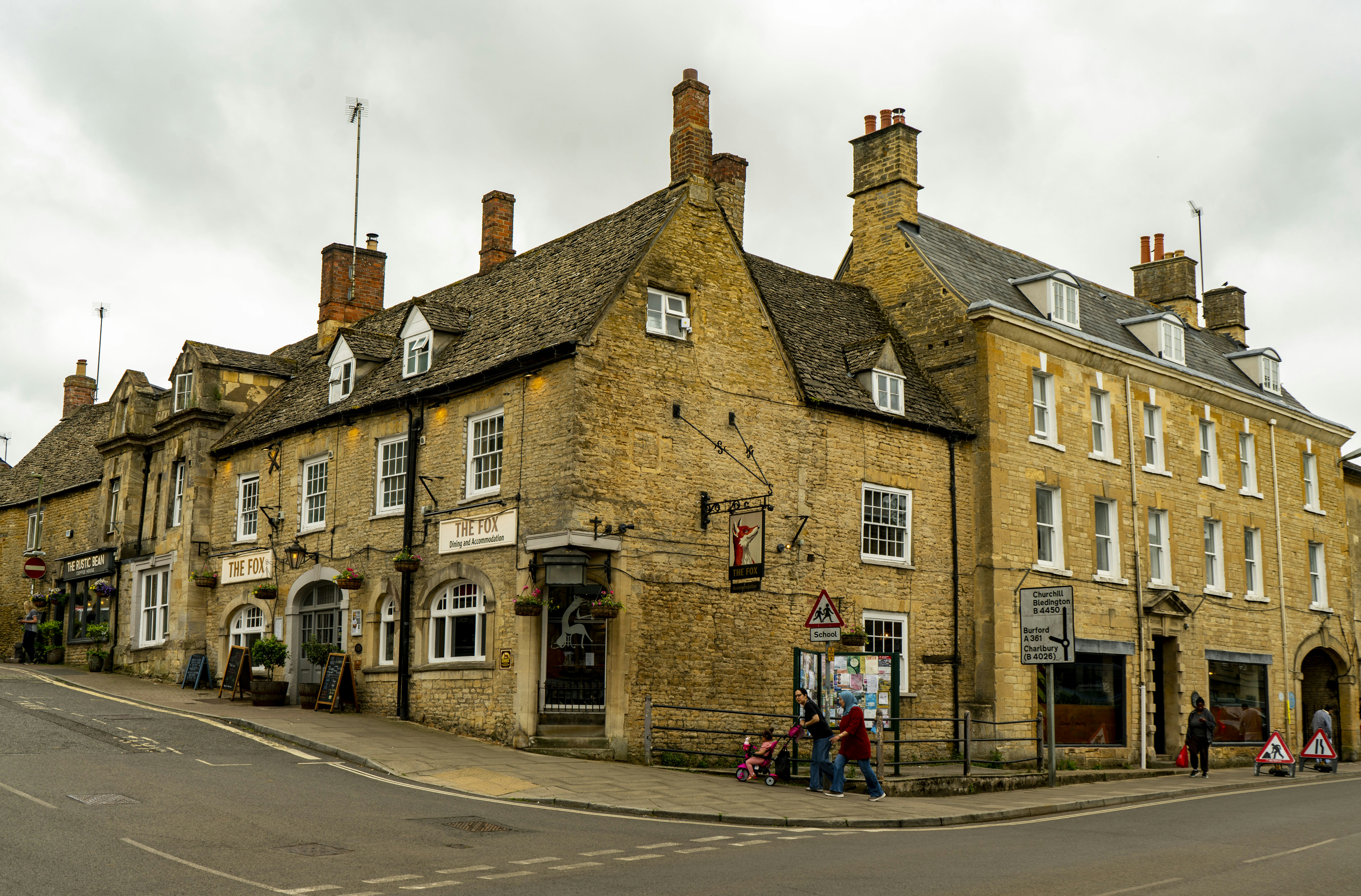 Stone storefronts line a quiet town corner with a pub and arched doorway; pedestrians and a person on a bicycle add life near the intersection.