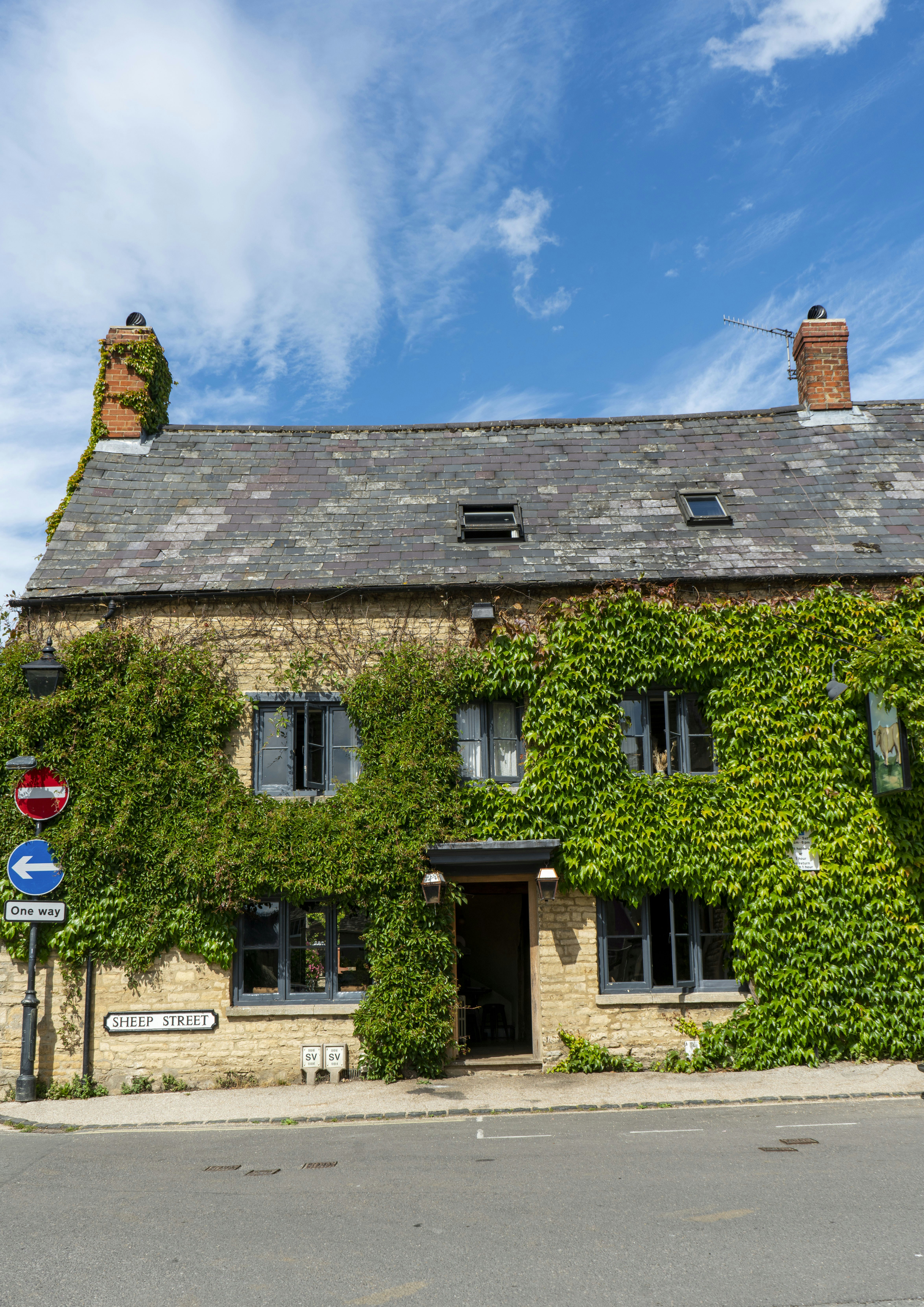 Stone cottage wrapped in ivy under a blue sky with a slate roof and chimneys, street-level scene with a doorway and roadside signs.