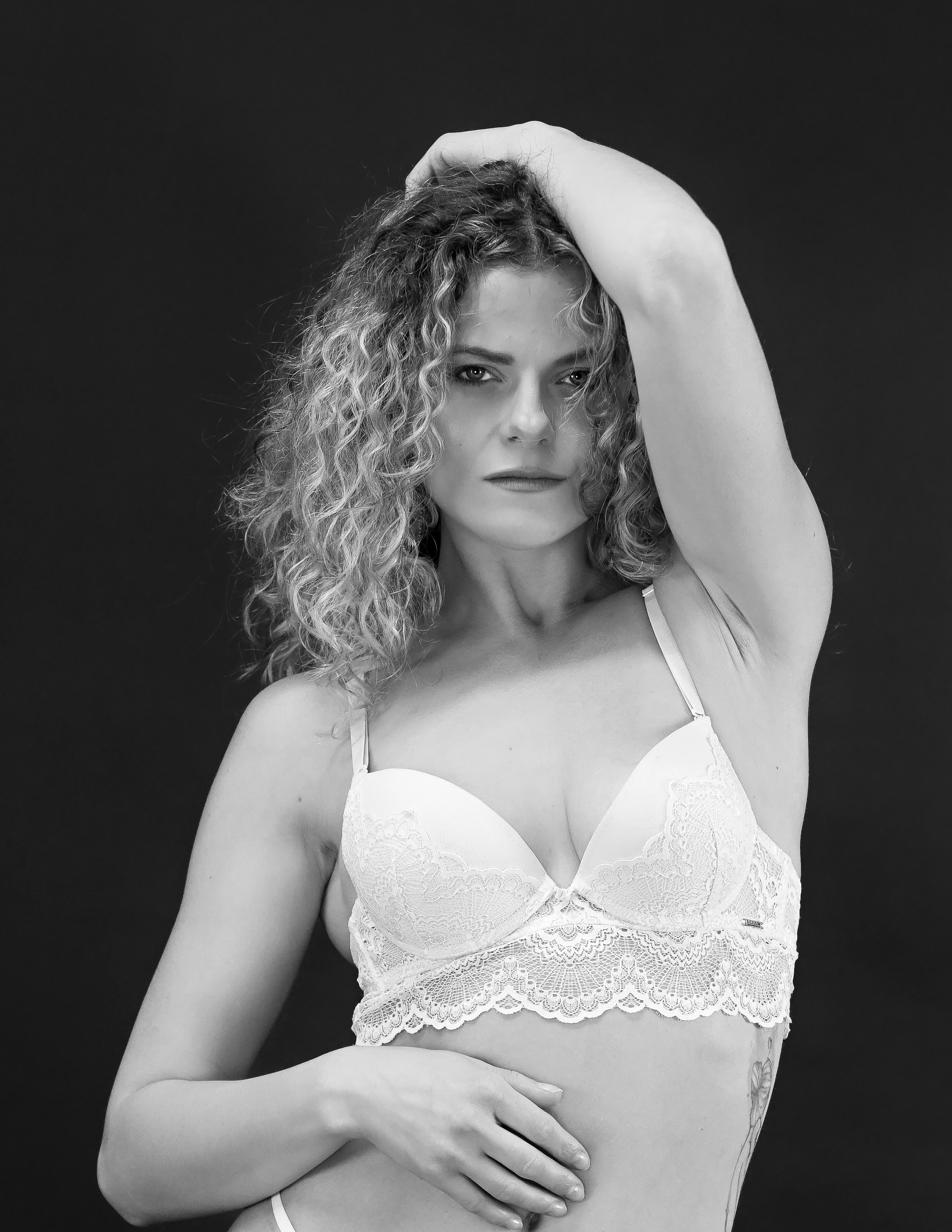High-contrast portrait of a woman in a lace bra, posed with one arm raised, against a dark backdrop. A minimalist studio shot emphasizing facial expression and texture of hair and fabric.