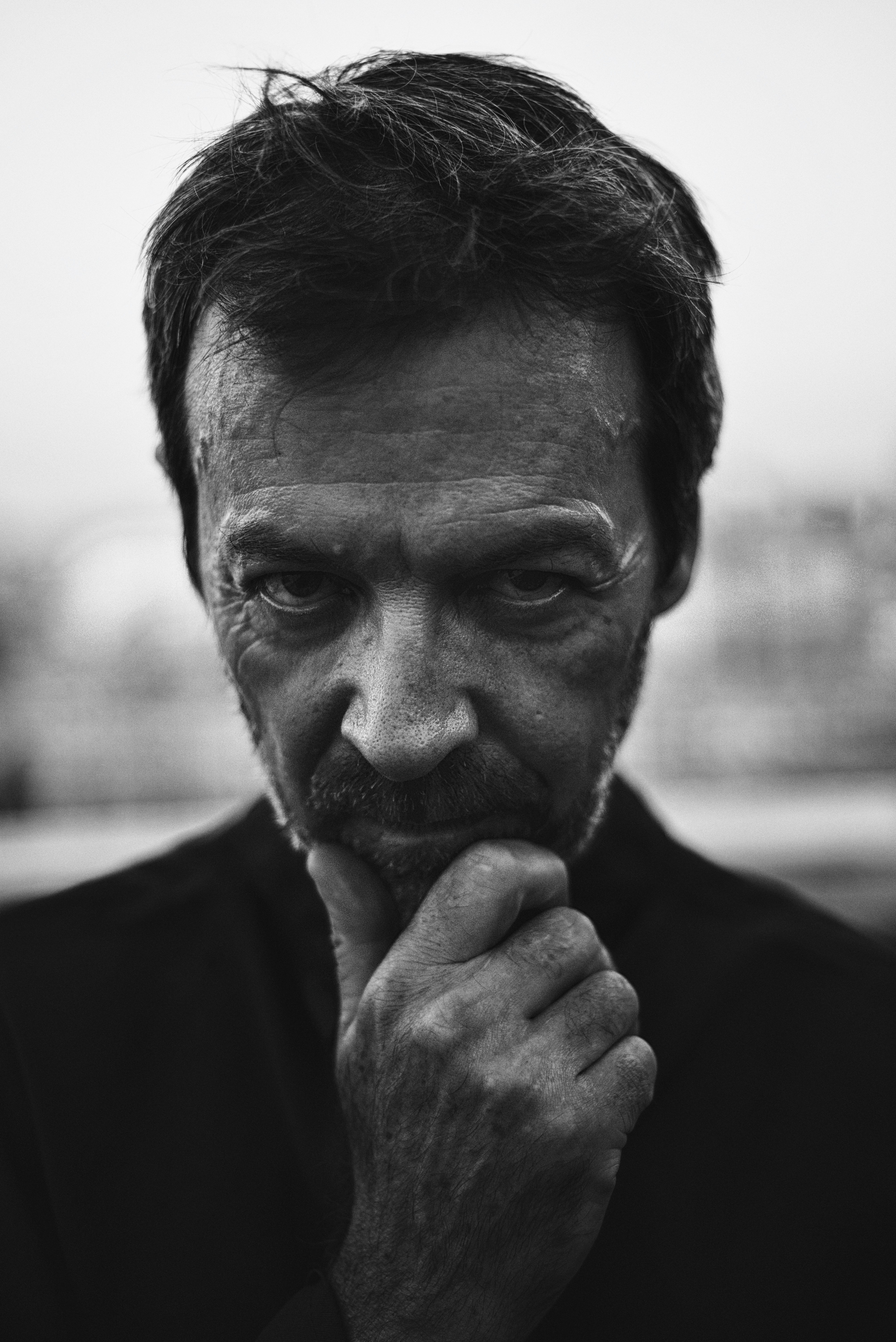 Close-up portrait of a man in black and white, fingers resting on his chin, shallow depth of field with a soft background.