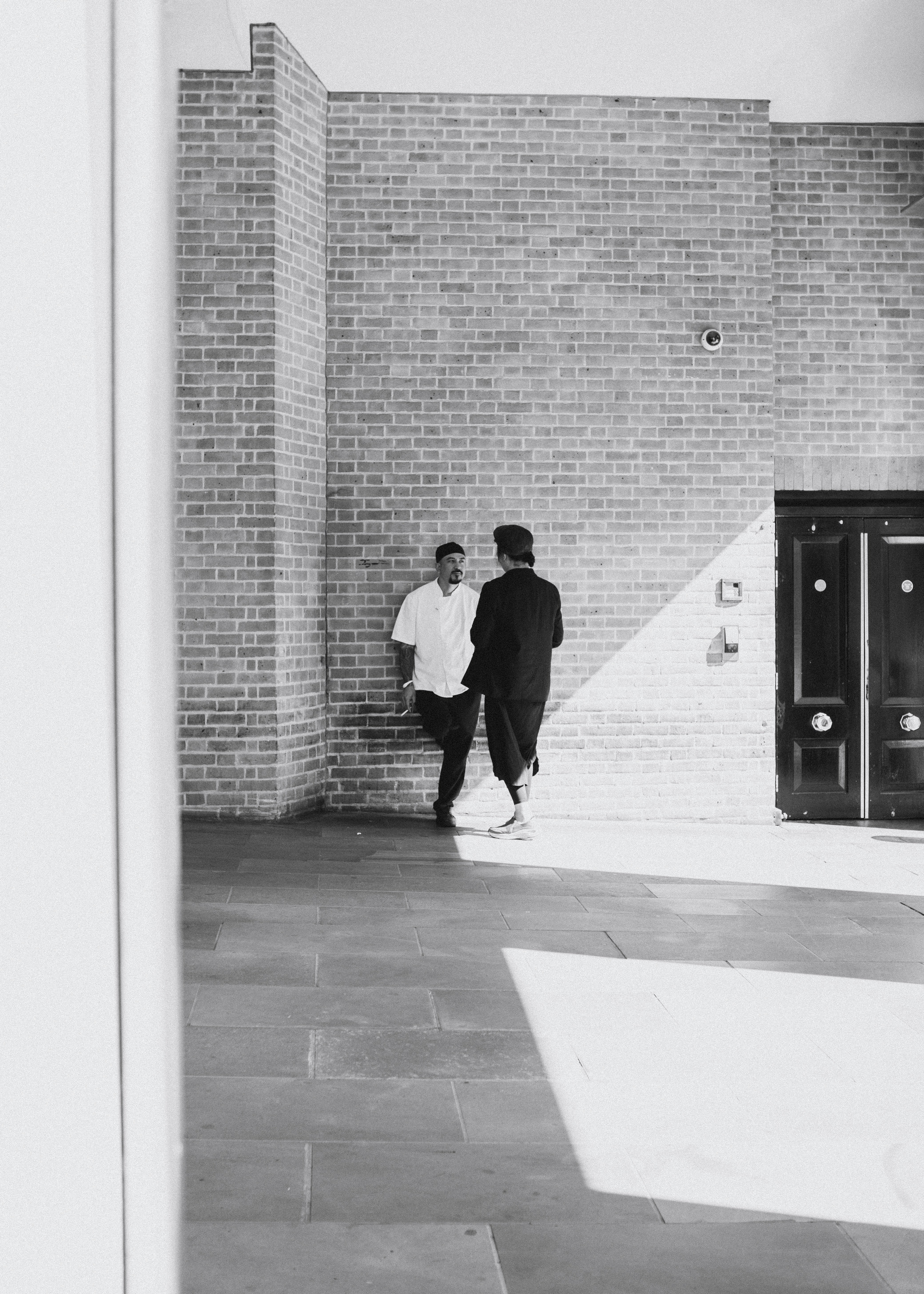 Two men stand near a brick wall in shaded daylight, one facing away in a dark jacket and the other in a light shirt; the scene is captured in black and white with architectural geometry and a large shadow polygon across the ground.