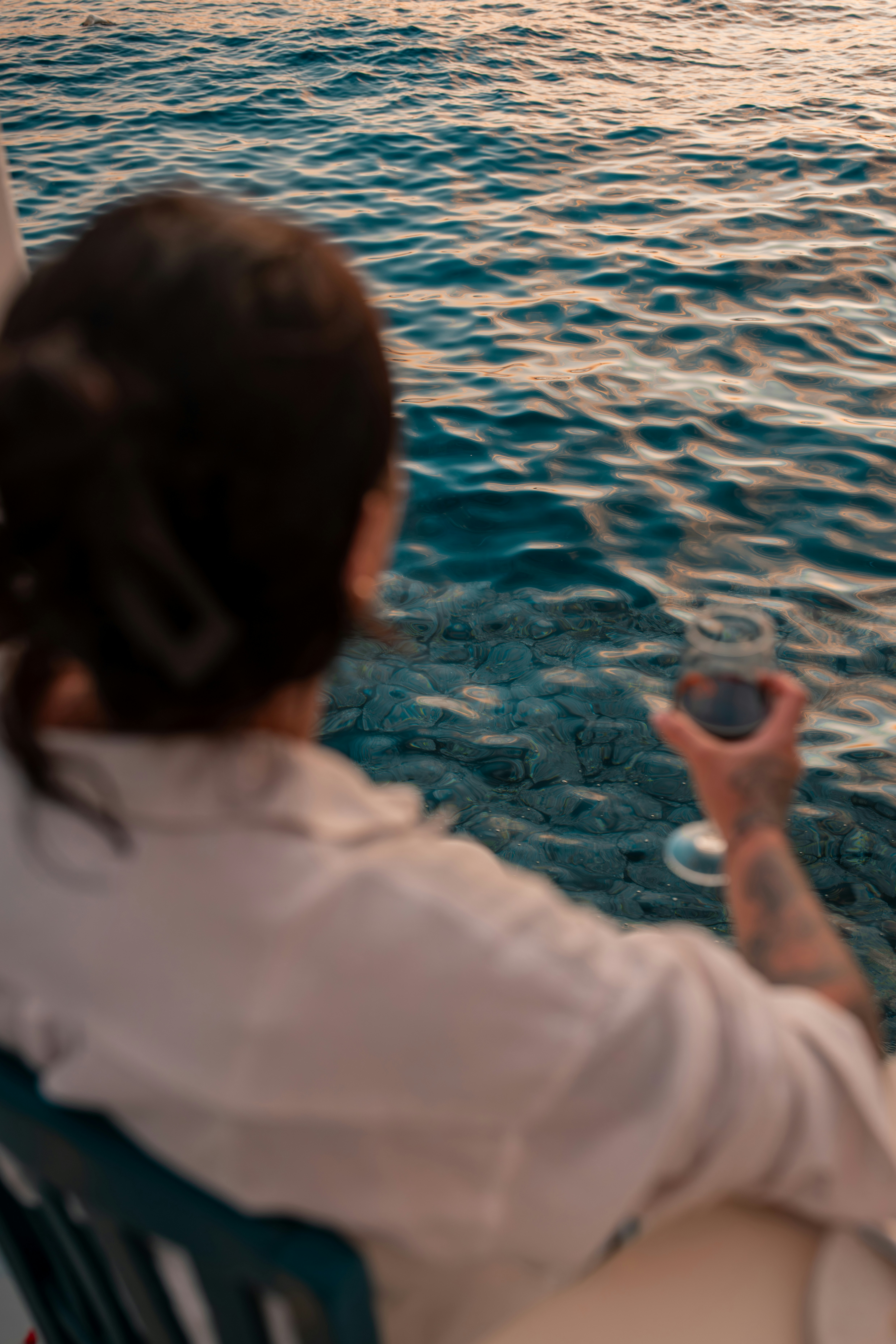 A person with a blurred back view lifts a glass over turquoise water, carving a calm, intimate moment near the edge of a boat.