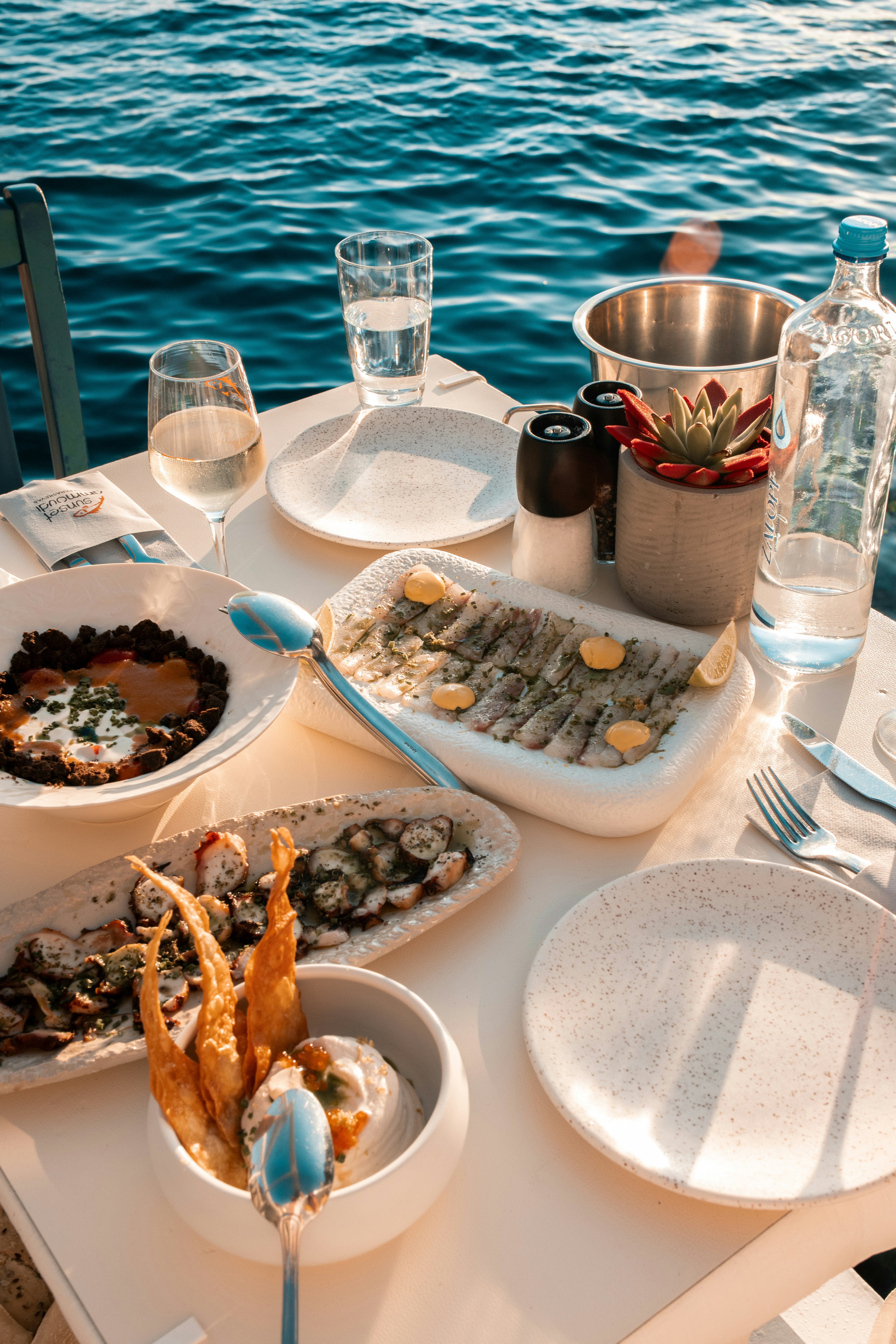 A sunlit table laden with seafood and drinks sits by sparkling blue sea, plates, utensils, and a succulent plant adding color to the scene. Food presentation and beverage details emphasize a relaxed coastal meal.