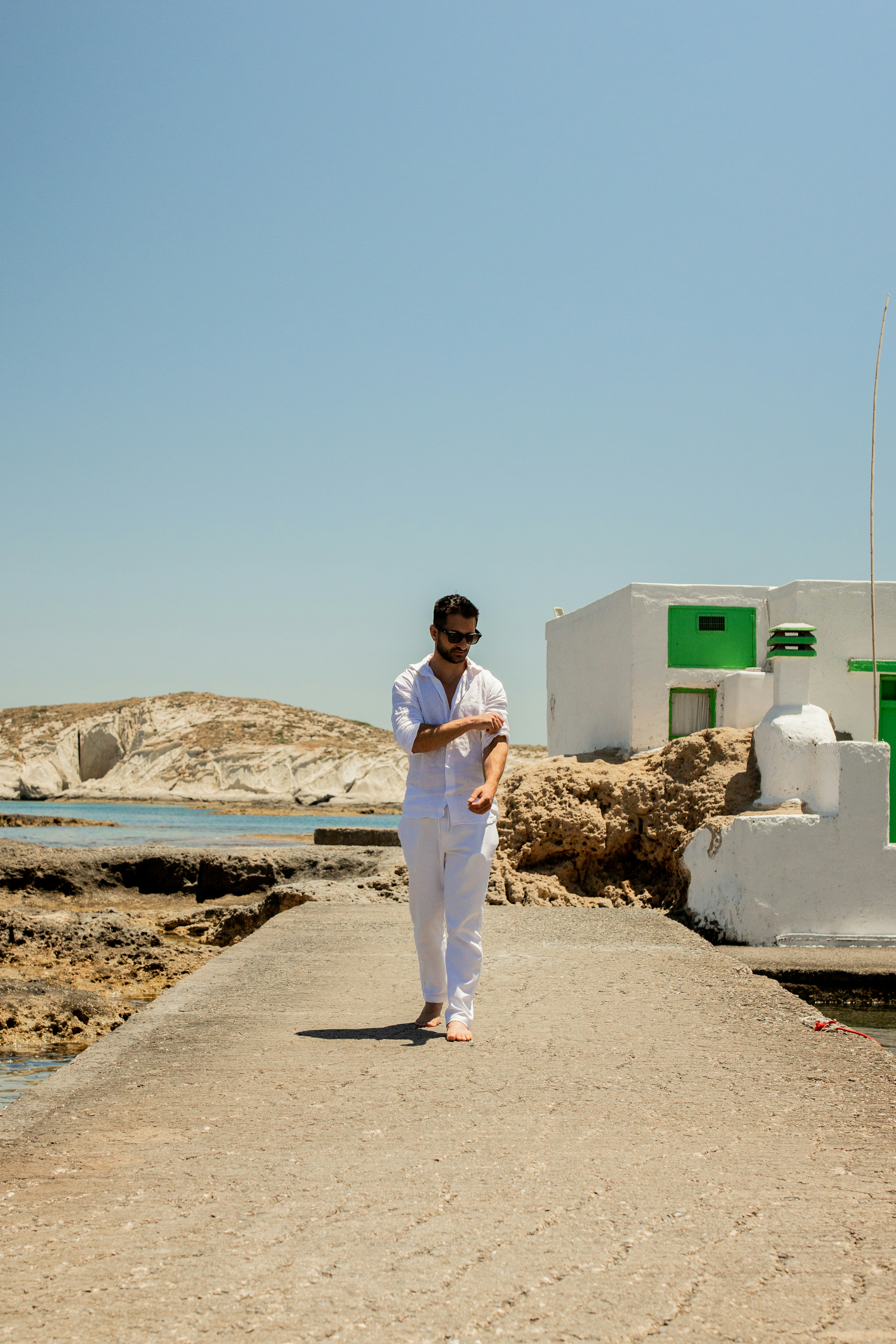 White-sailed charm: a man in white walks the coastal jetty under a clear sky