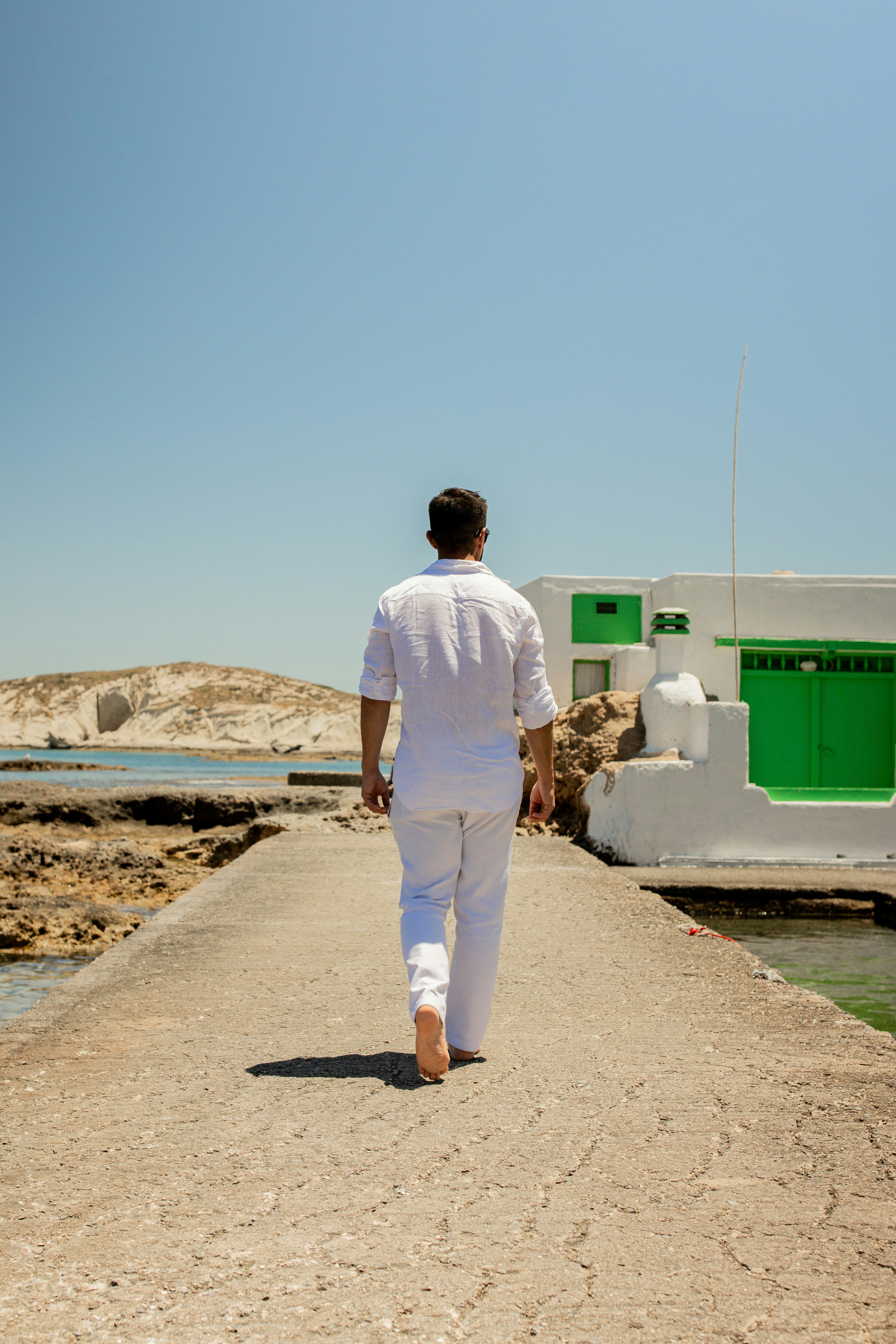 White-sleeved traveler walks along a sunlit sea pier toward a white-green waterfront building