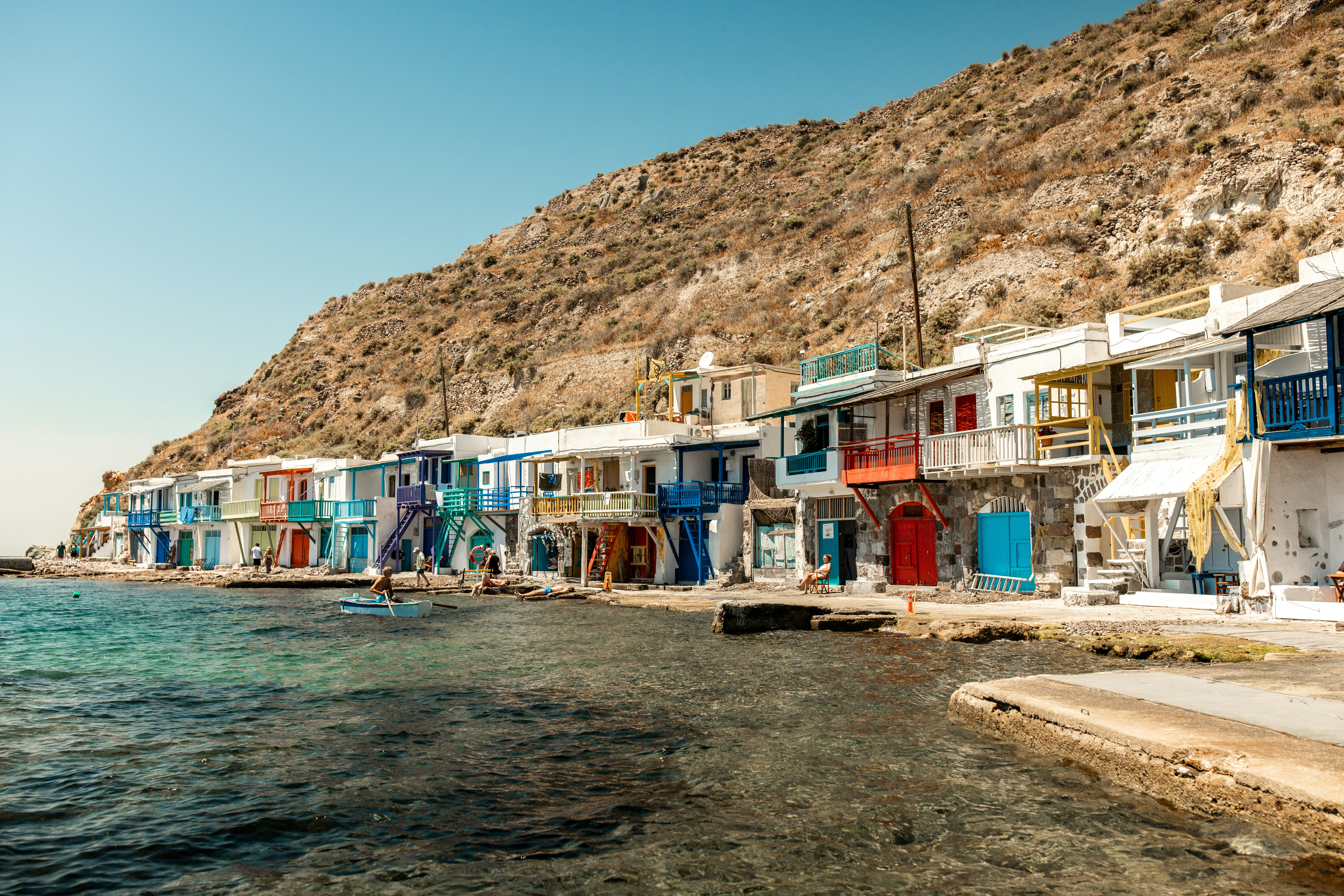 Colorful seaside huts line a rocky shoreline under a sunlit hillside