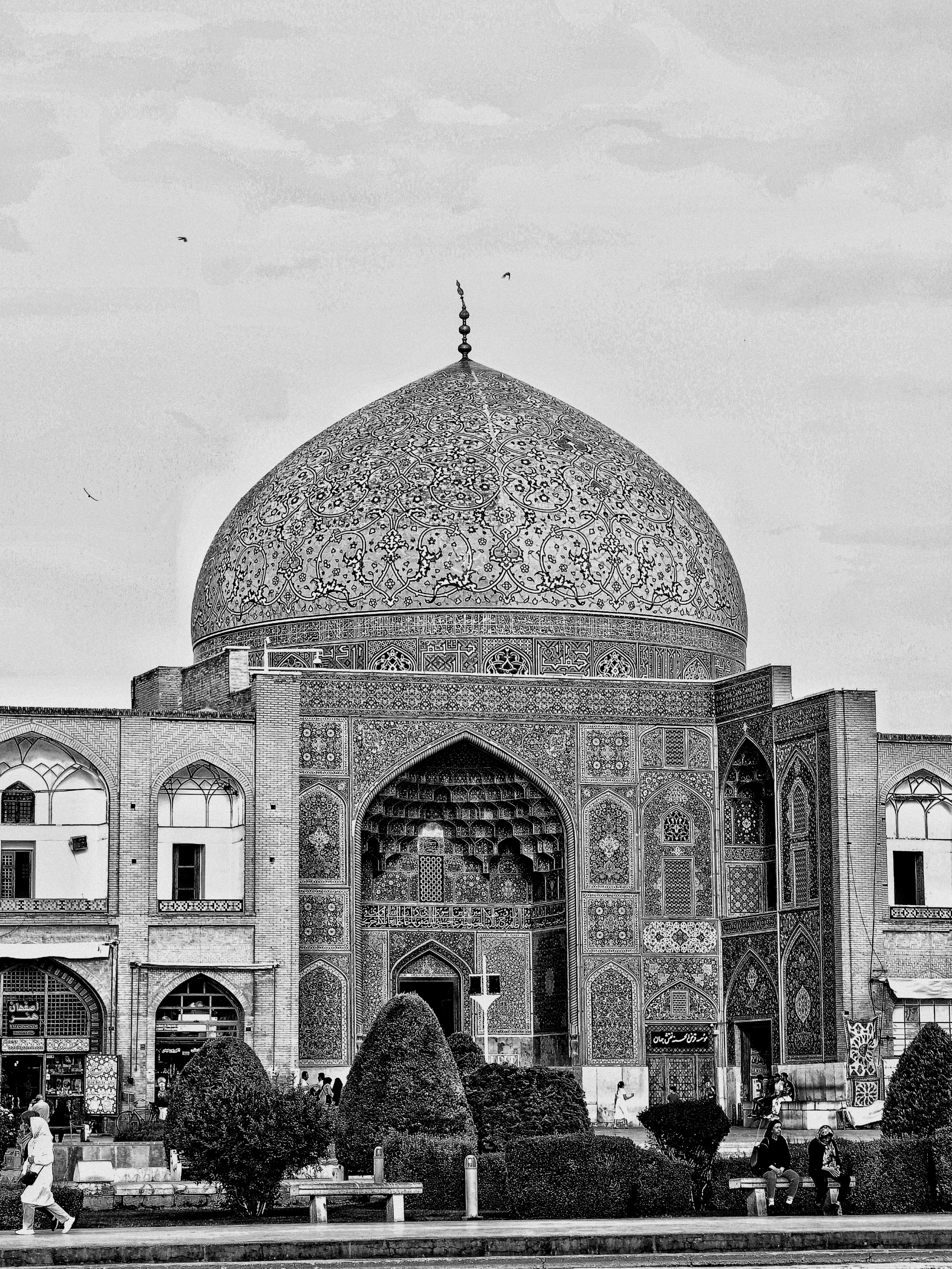 Intricately patterned mausoleum facade with a large domed top, flanked by arches and manicured shrubs in a formal garden.