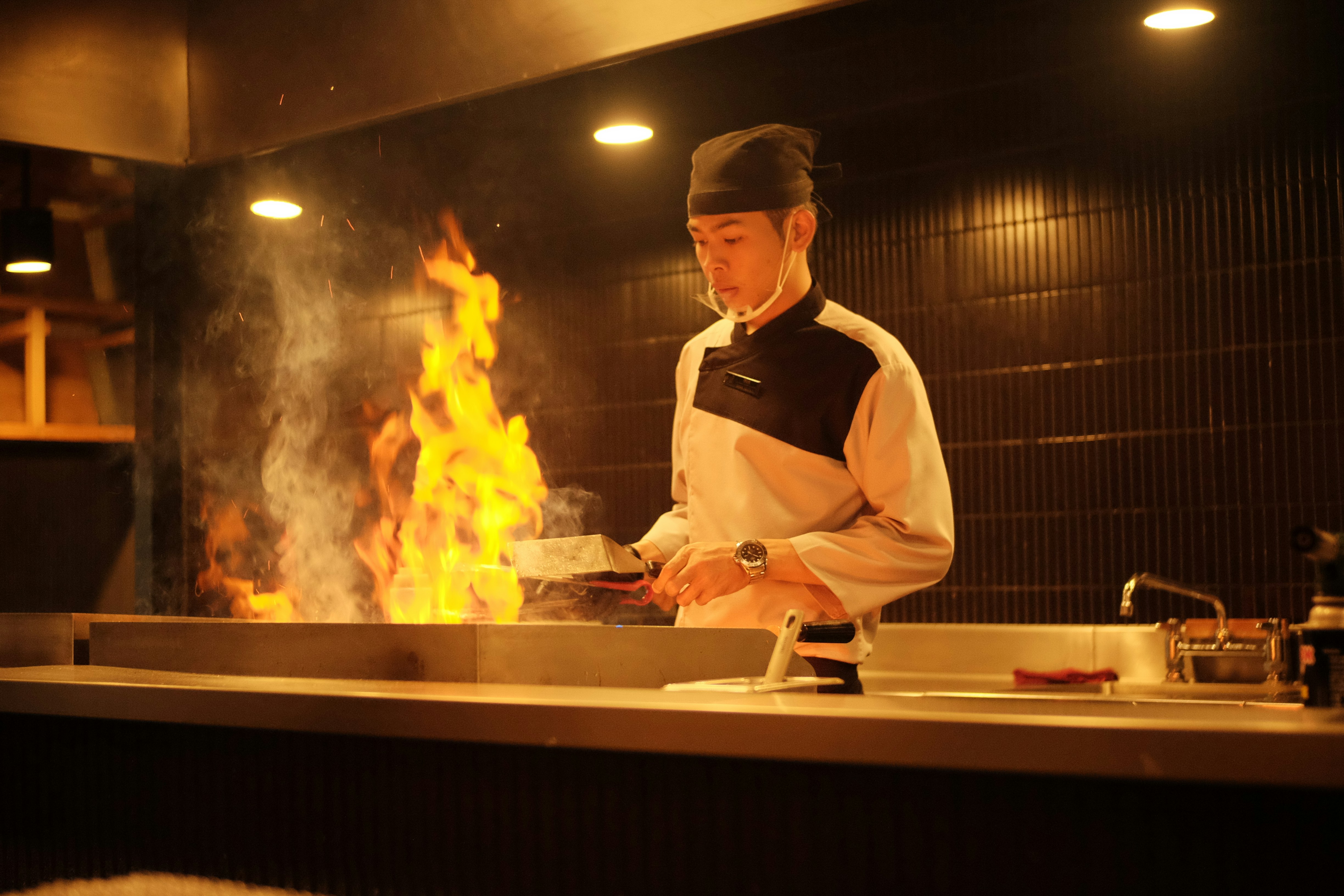 A chef stands at a sizzling open flame station, concentrating on a task with firelight highlighting the workspace in a dim kitchen ambiance.