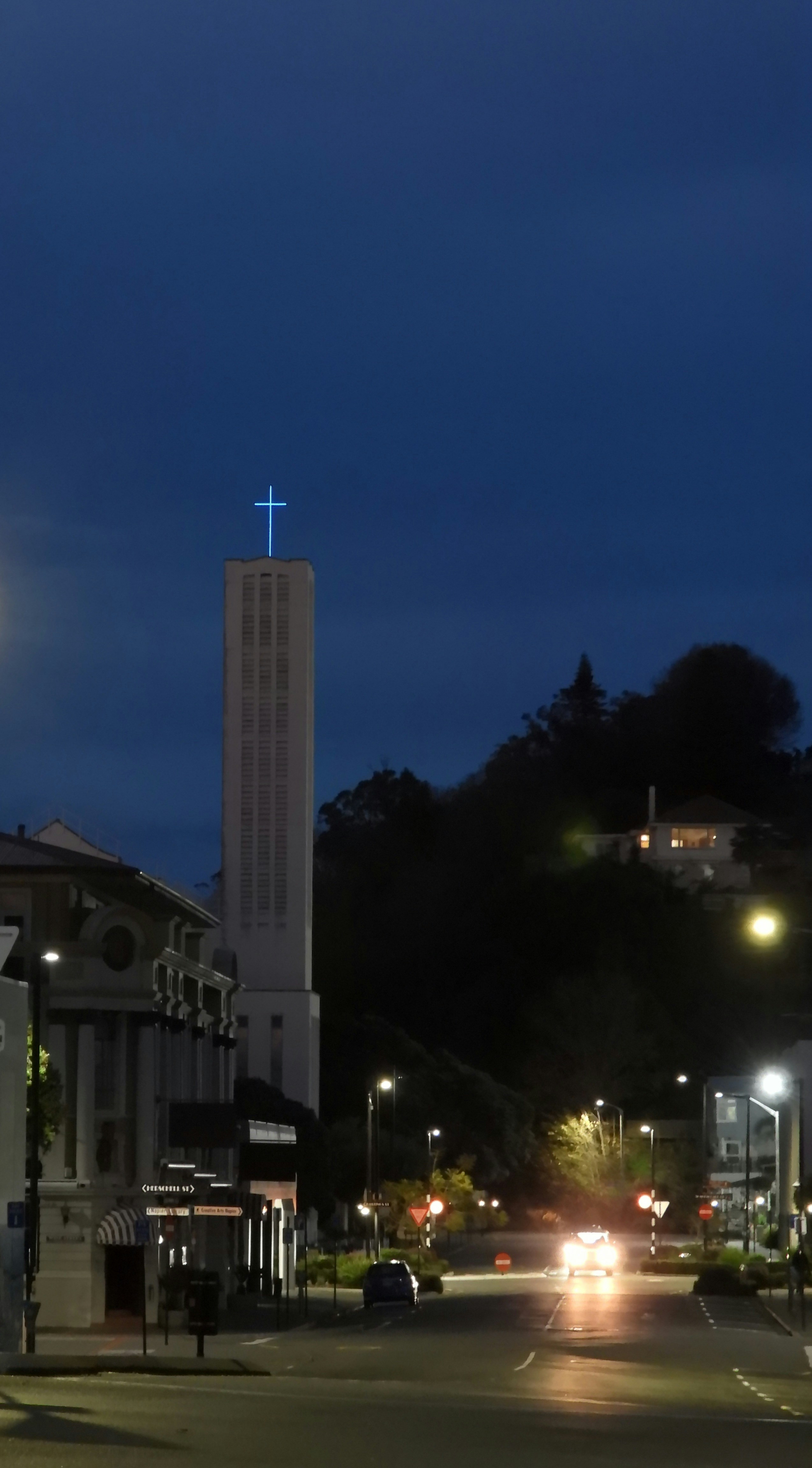 A quiet urban street at dusk with a tall church tower and illuminated cross rising above storefronts and trees, casting gentle street glow.