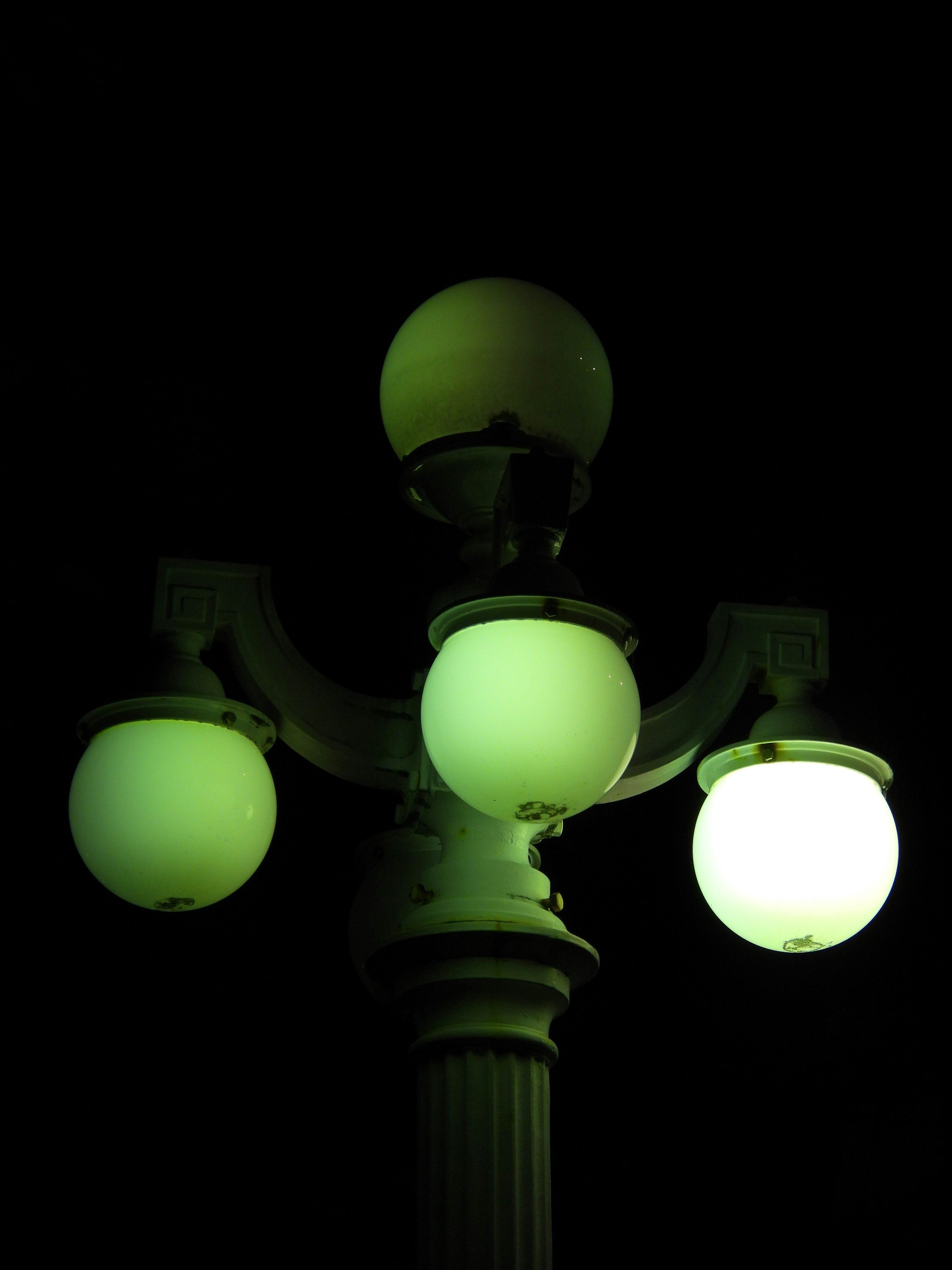 Green-lit globe lamps illuminate a classic lamppost against a dark night, highlighting vintage architecture and moody illumination. This can be a part of architectural or urban-night photography.