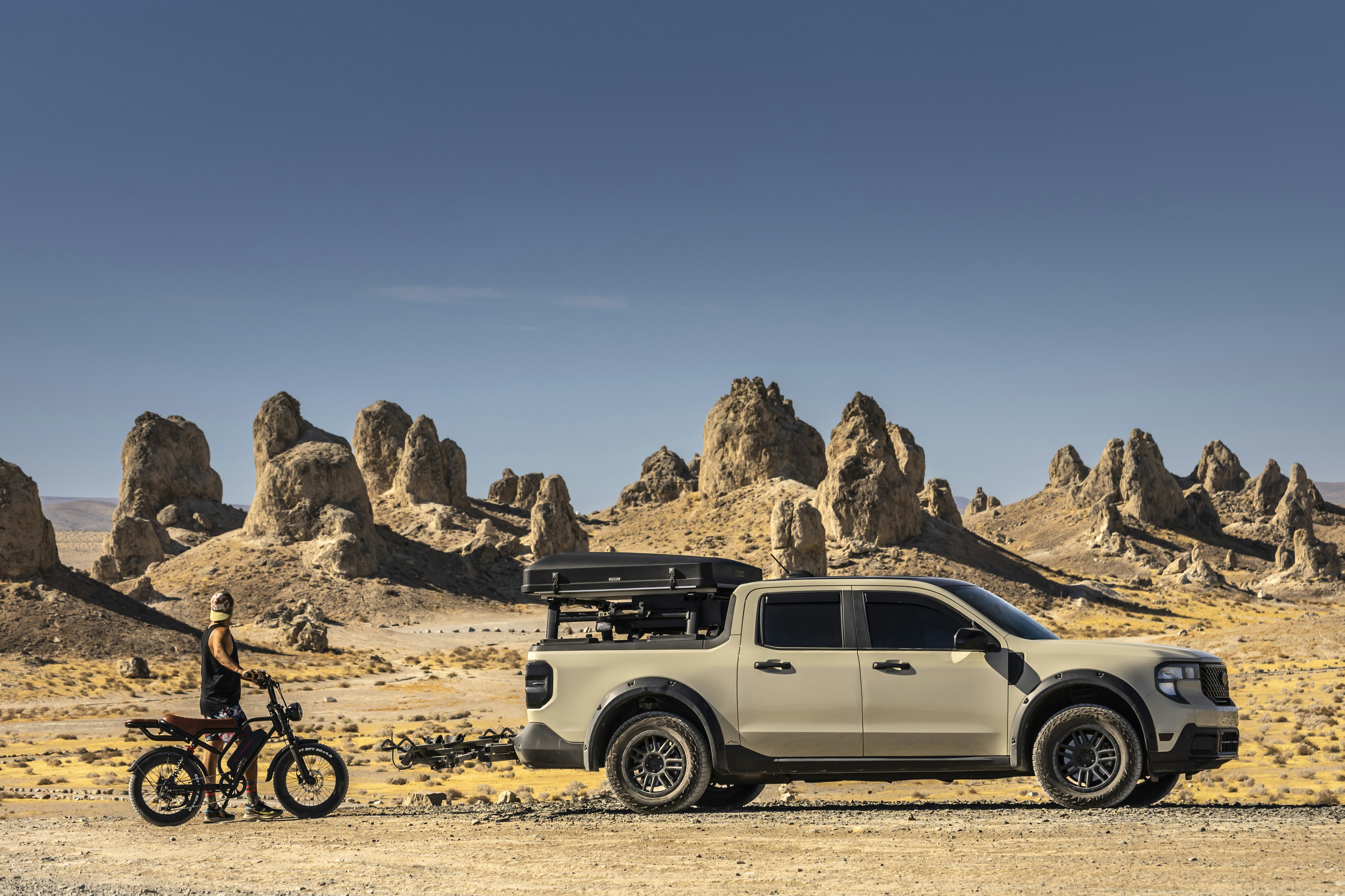 Beige SUV and a cyclist stand by a rugged desert landscape with jagged rock formations under a clear blue sky.