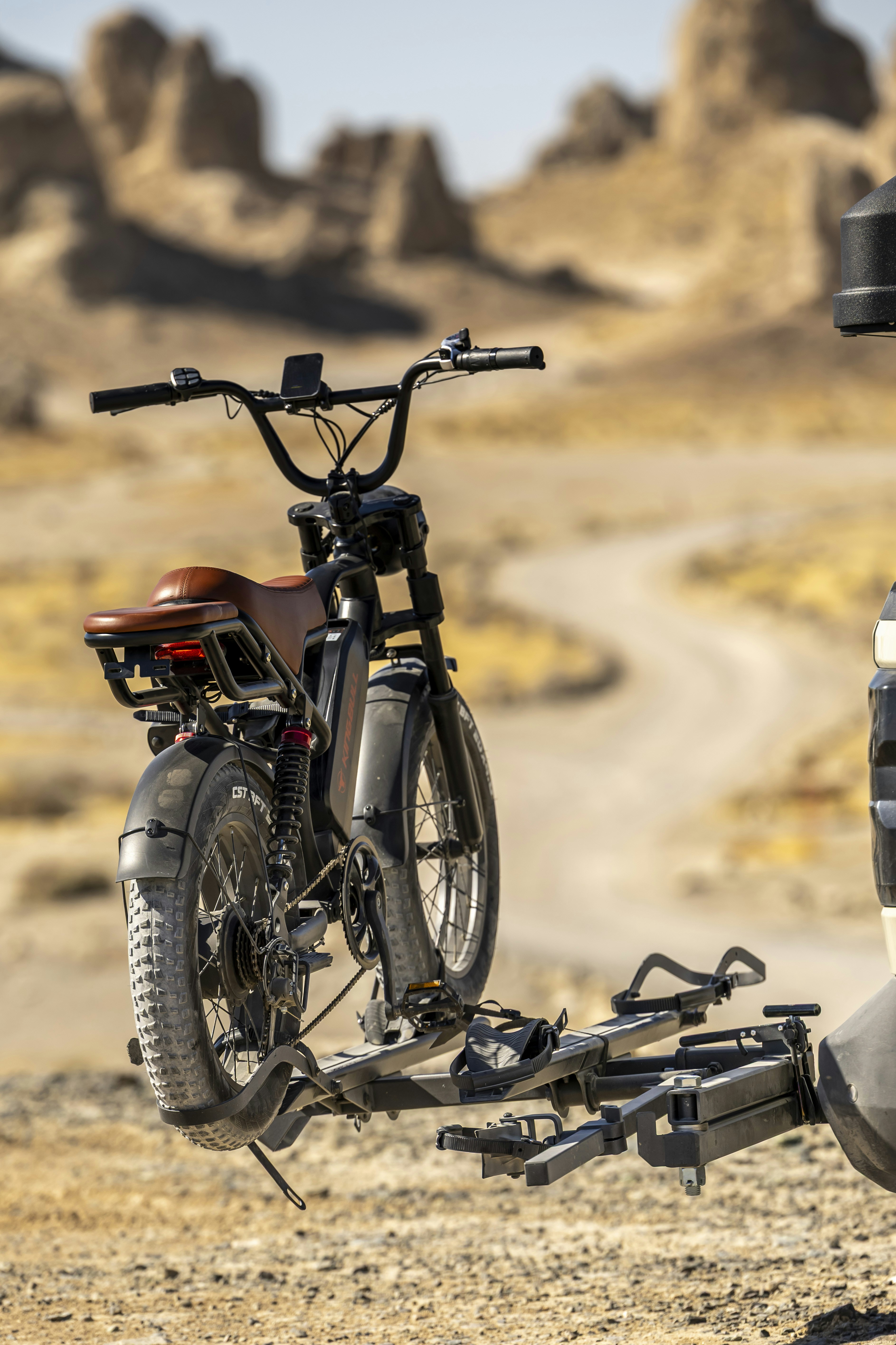 A rugged bike sits on a trailer hitched to a vehicle in a desert landscape, with rocky formations blurred in the background. A portable bike stand and the trailer hitch are visible, capturing a moment of outdoor adventure and transport.
