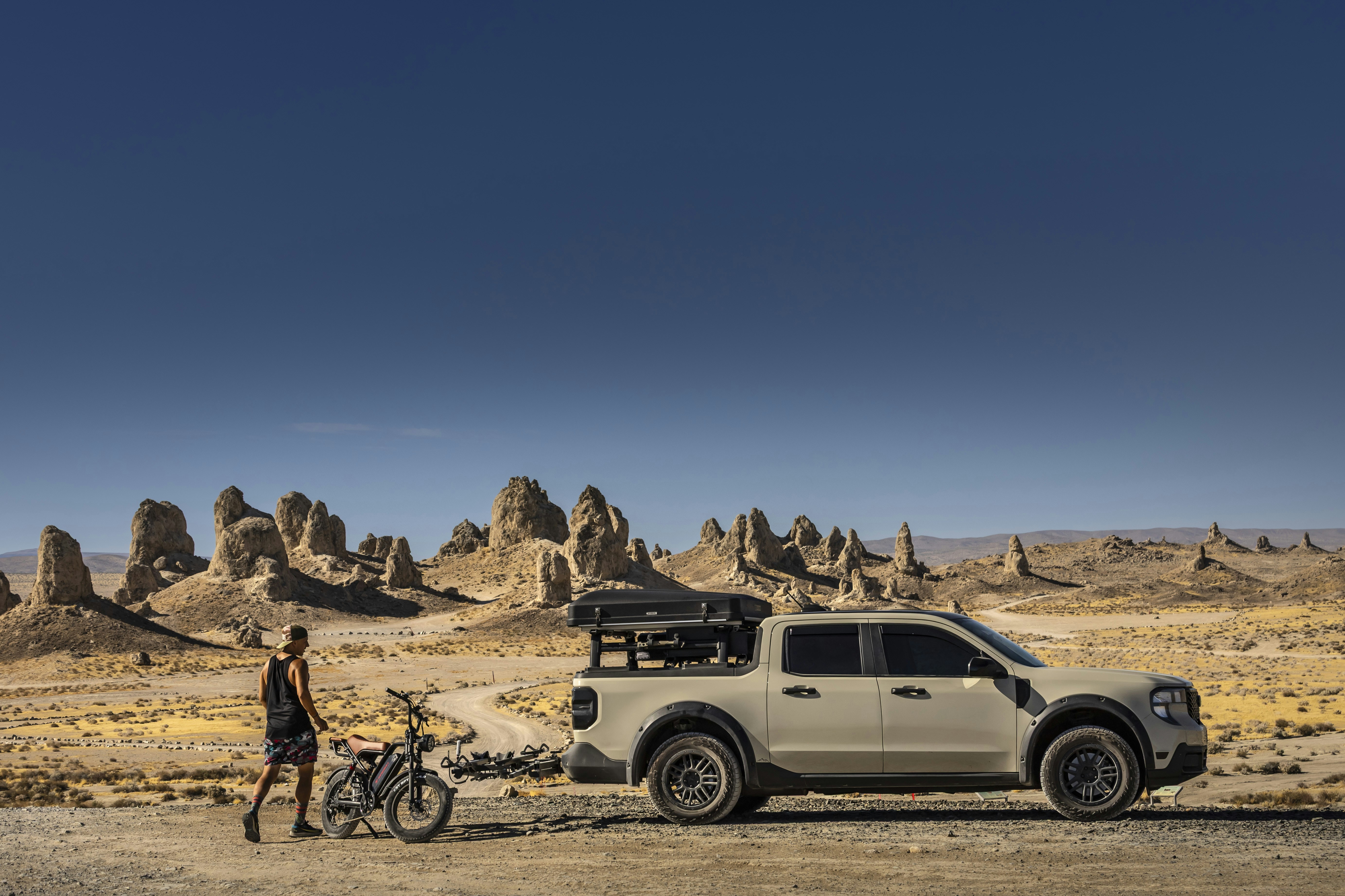 Desert landscape with a man, a bicycle, and a pickup parked under a clear blue sky; a rugged formation of rocky spires lines the horizon. A purpose-built expedition scene.