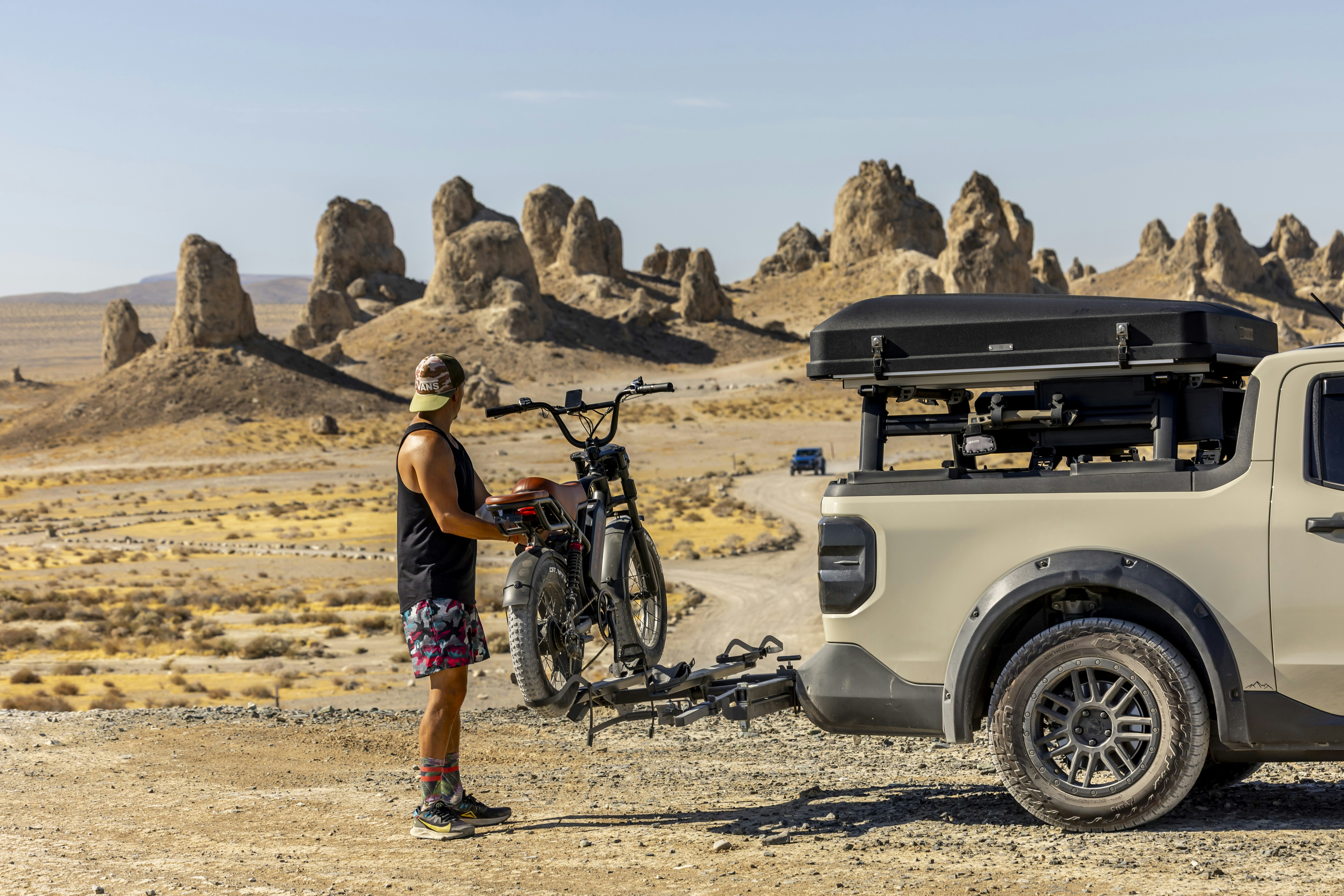 Rugged traveler with a muscle bike and a hitch-mounted trailer works beside a pickup in a sunlit desert, with eroded rock formations in the distance.