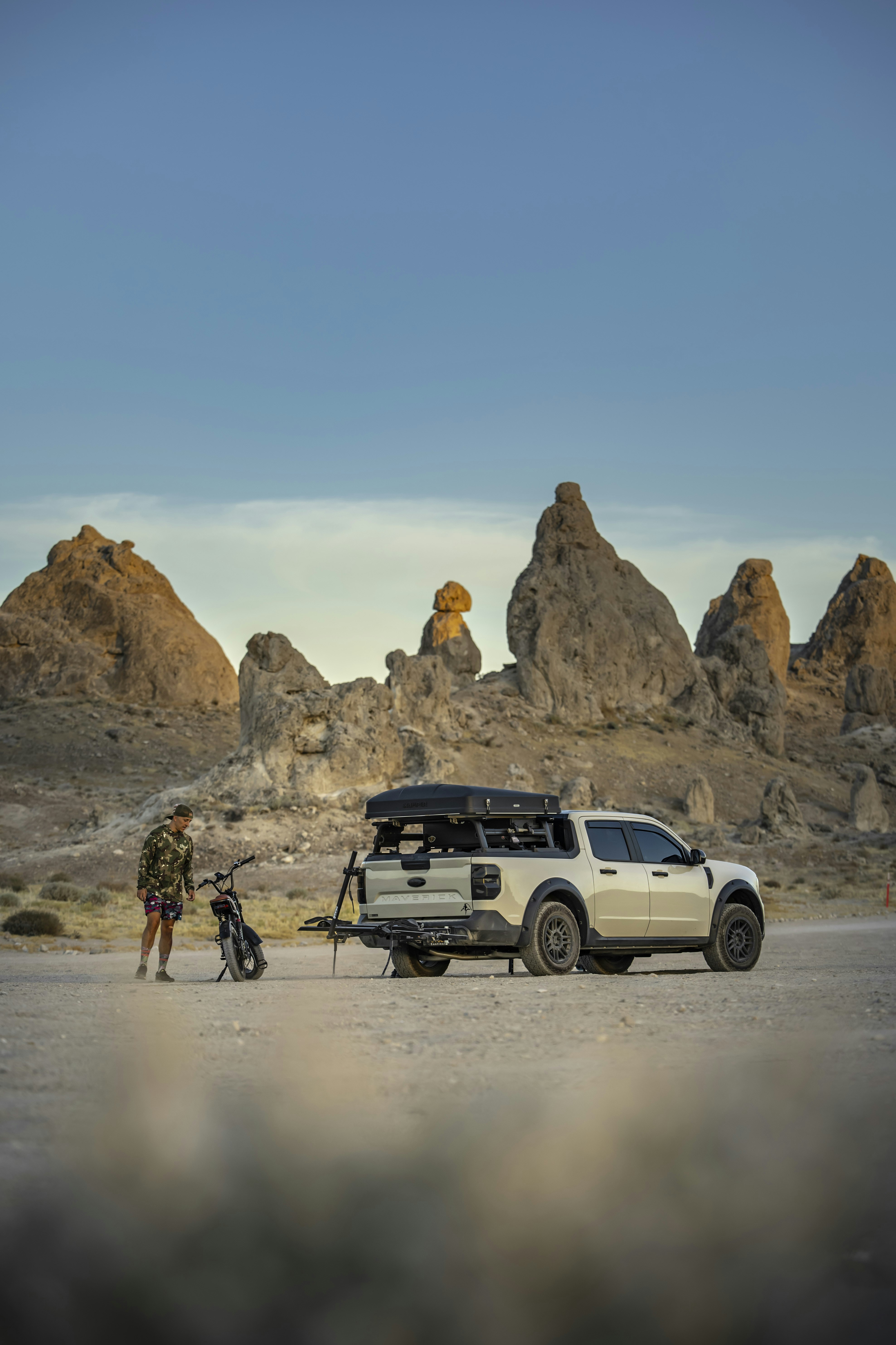 Off-road scene in a desert basin with a rugged SUV and a rider with a bike, framed by jagged rock formations under a clear blue sky. A stationary setup emphasizing exploration gear and landscape scale.