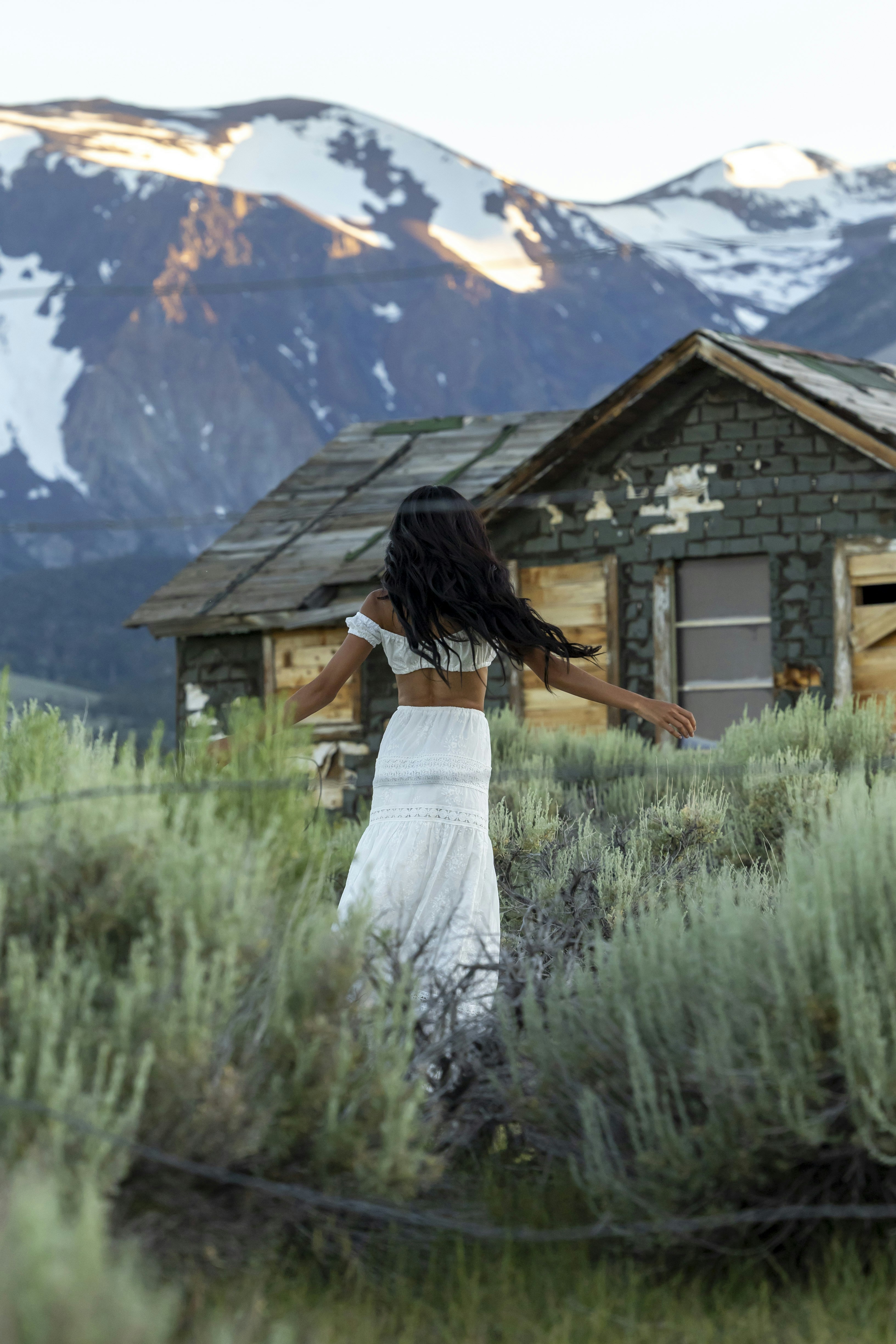 A woman in a white dress walks toward a weathered cabin with snow-capped mountains in the background, sagebrush foreground, soft natural light. A serene, bucolic scene with rustic textures.