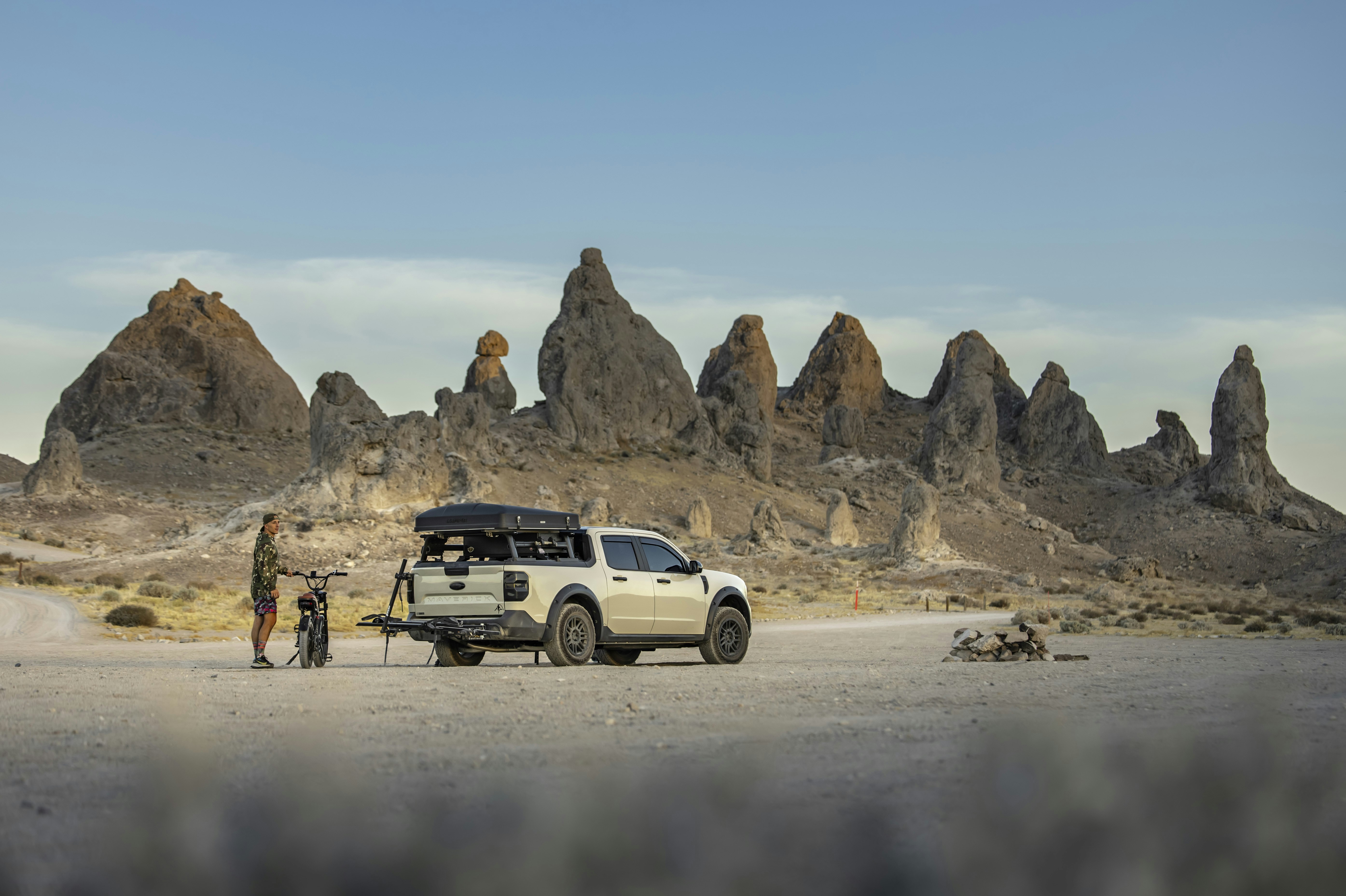Off-road SUV parked on pale desert floor with two explorers beside a toolkit, jagged basalt spires rising in the background under a clear blue sky.