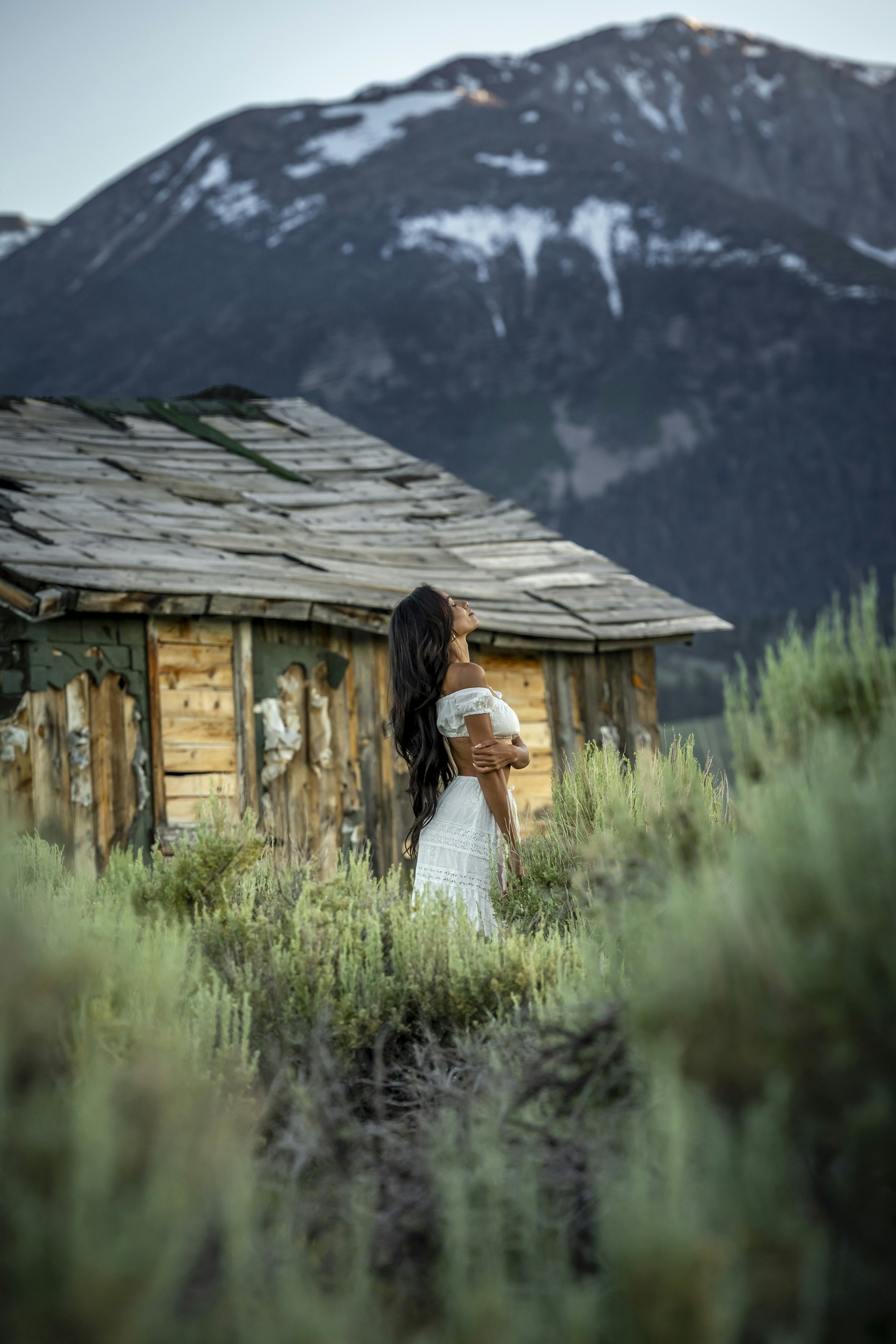 A woman in a white dress stands beside a weathered wooden cabin in a meadow with a snow-capped mountain backdrop. The scene emphasizes solitude and rustic beauty.