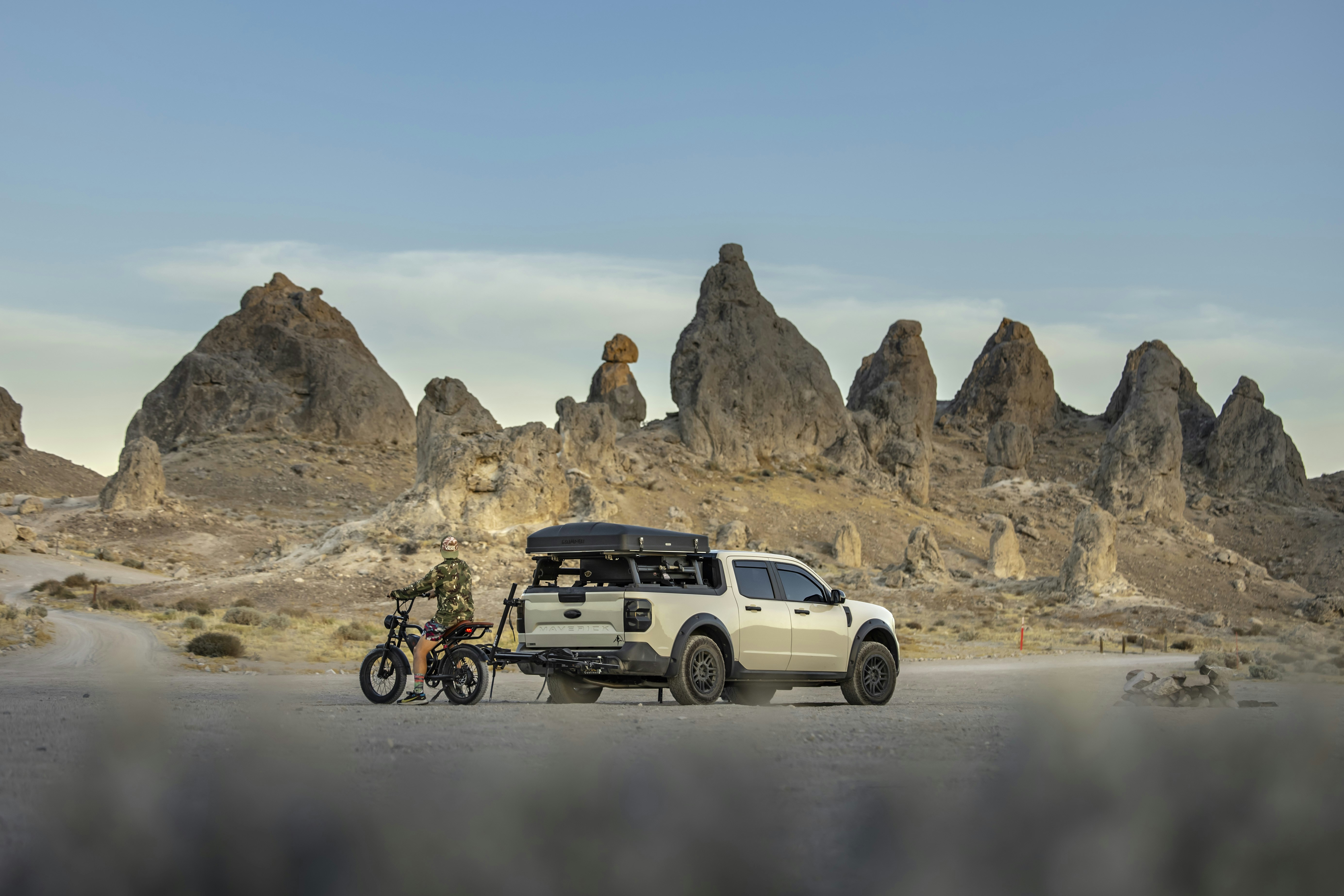 Off-road pickup with bike in tow rests on a gravel foreground beneath jagged rock formations; rugged landscape and distant blue sky establish a remote expedition scene.