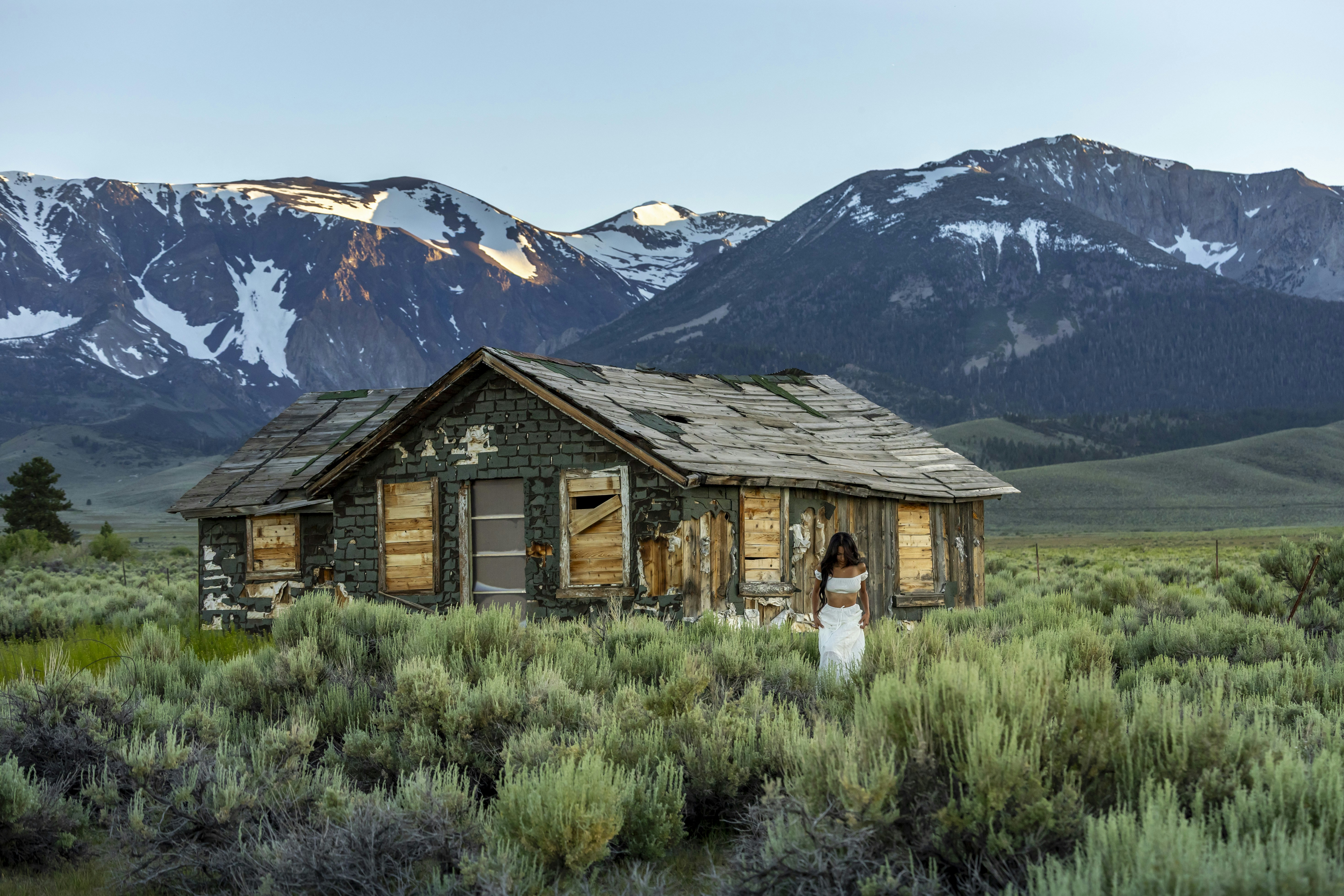 Weathered cabin sits among sagebrush with snow-capped mountains in the background; a person in white stands near the doorway, adding a human element to the desolate landscape.