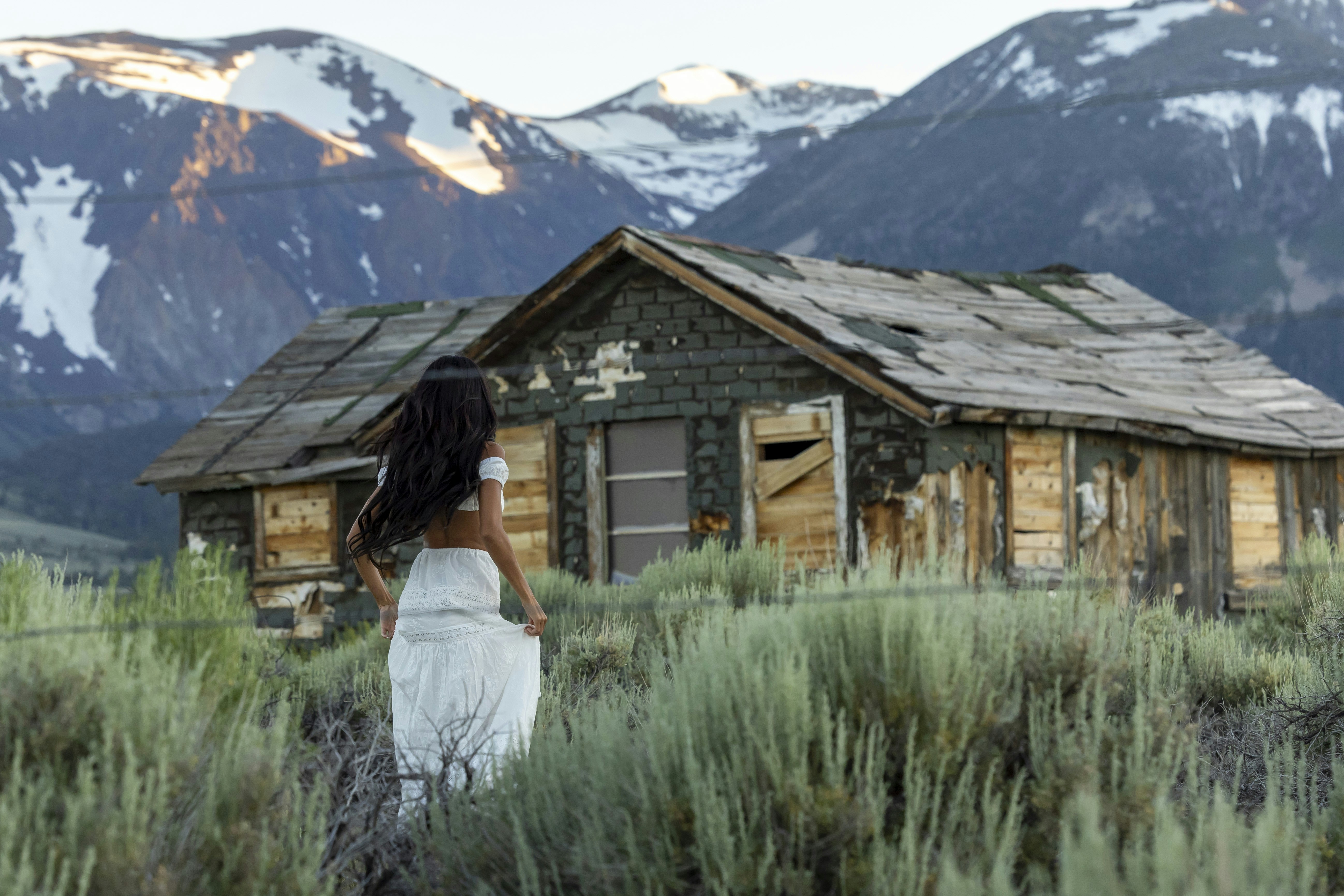A woman in a white dress walks toward a weathered, wooden cabin set against snow-capped mountains and scrubby foreground vegetation.